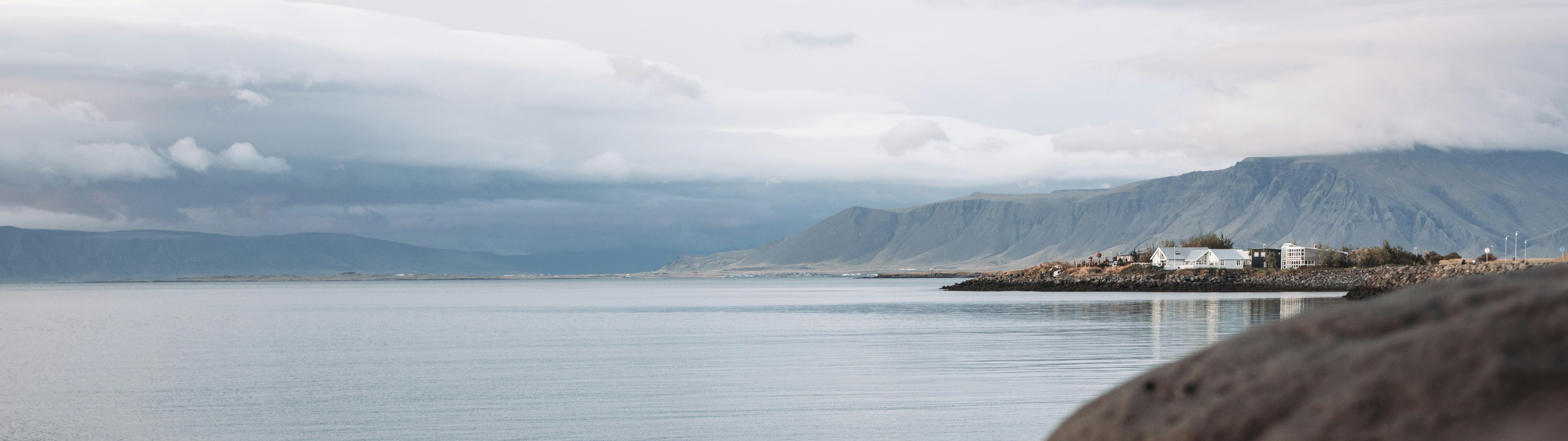 Calm waters stretch towards distant mountains under a cloudy sky, with a small coastal settlement on the right.