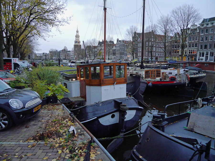 Moored boats line a canal in a European city, with historic buildings and a church tower in the background. Autumn leaves cover the cobblestone path along the water.