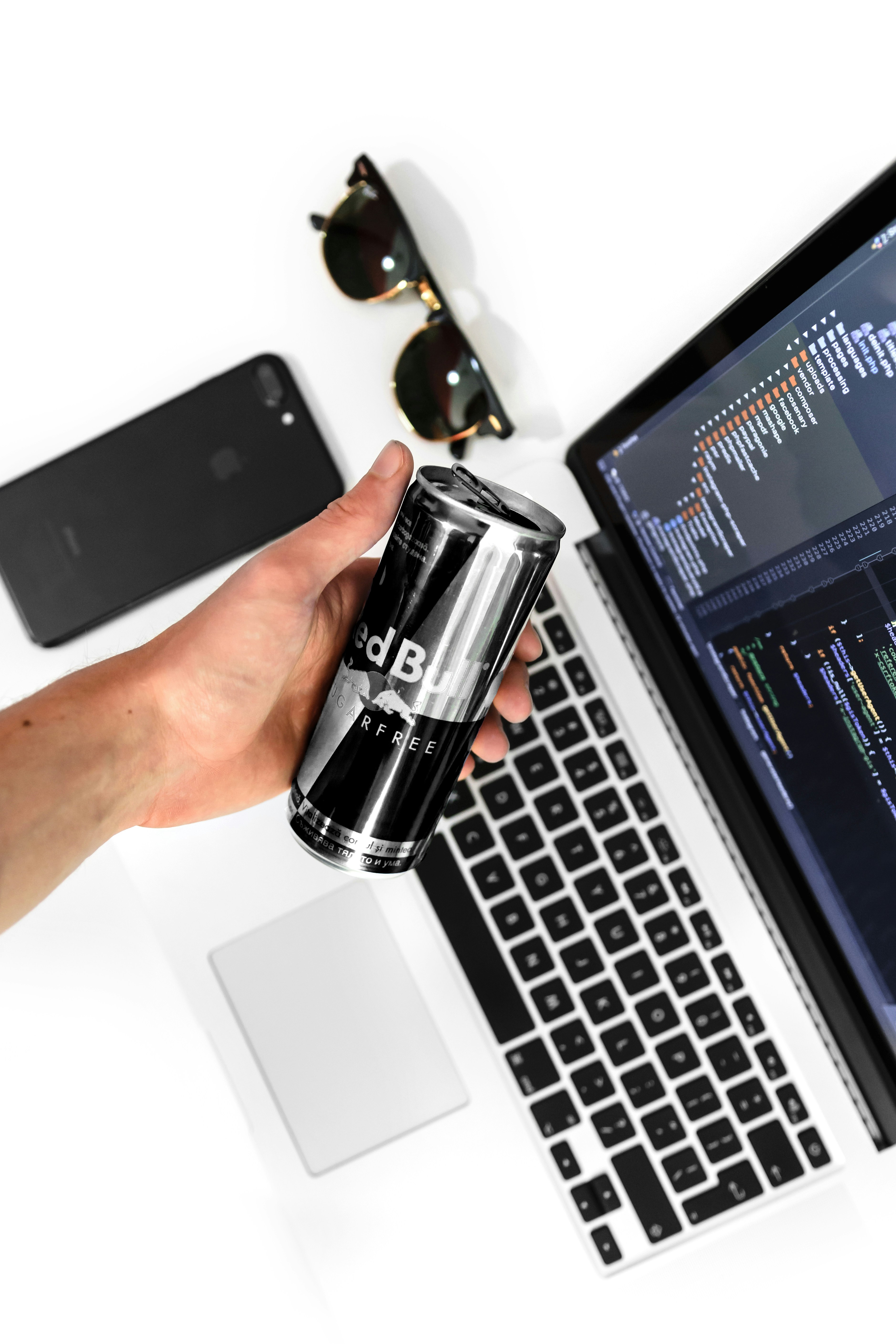 Hand holding a Black can next to a laptop displaying code, with a smartphone and sunglasses in the soft, bright background.