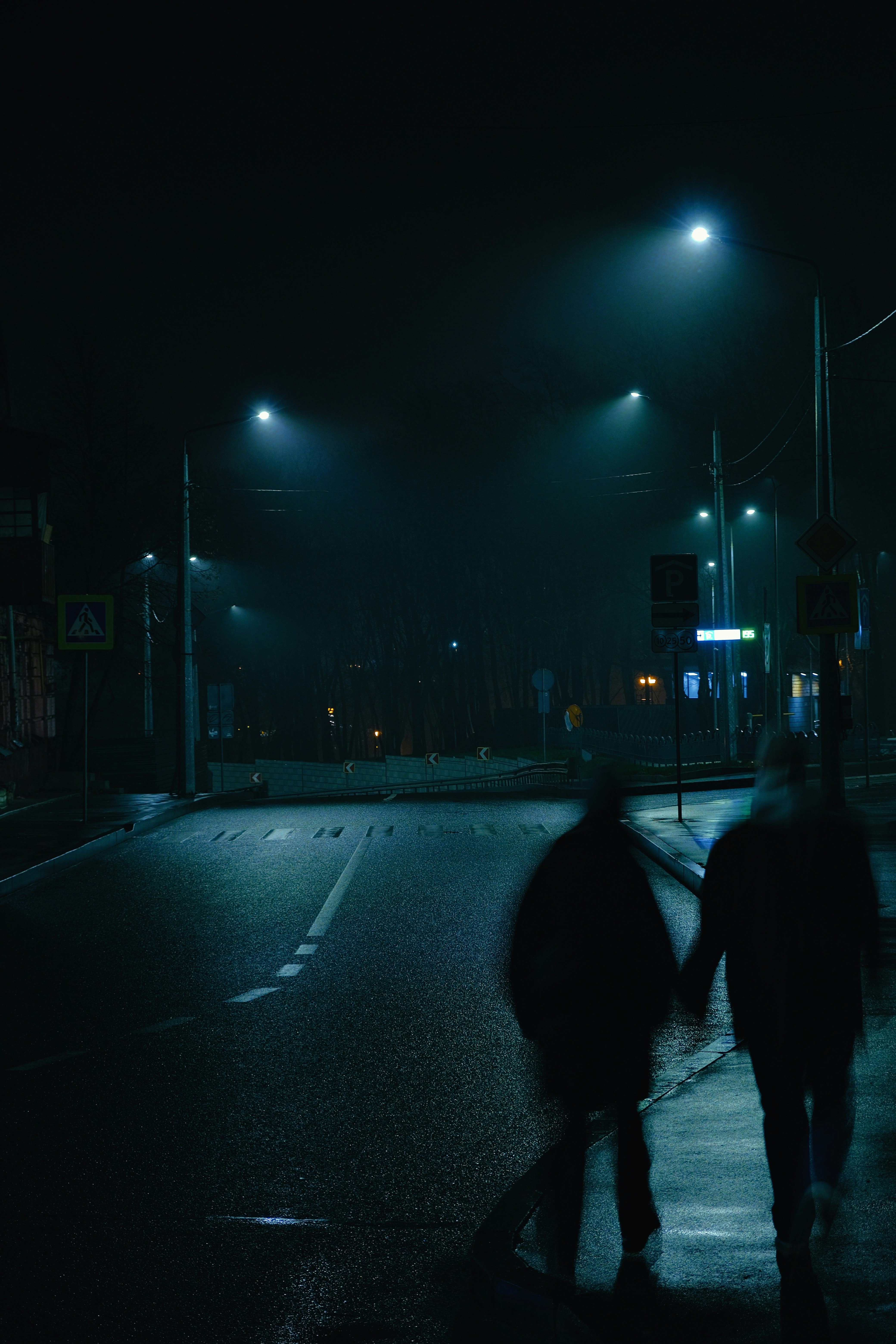 Silhouetted figures walking hand in hand on a deserted street illuminated by soft, hazy streetlights in a foggy night scene.
