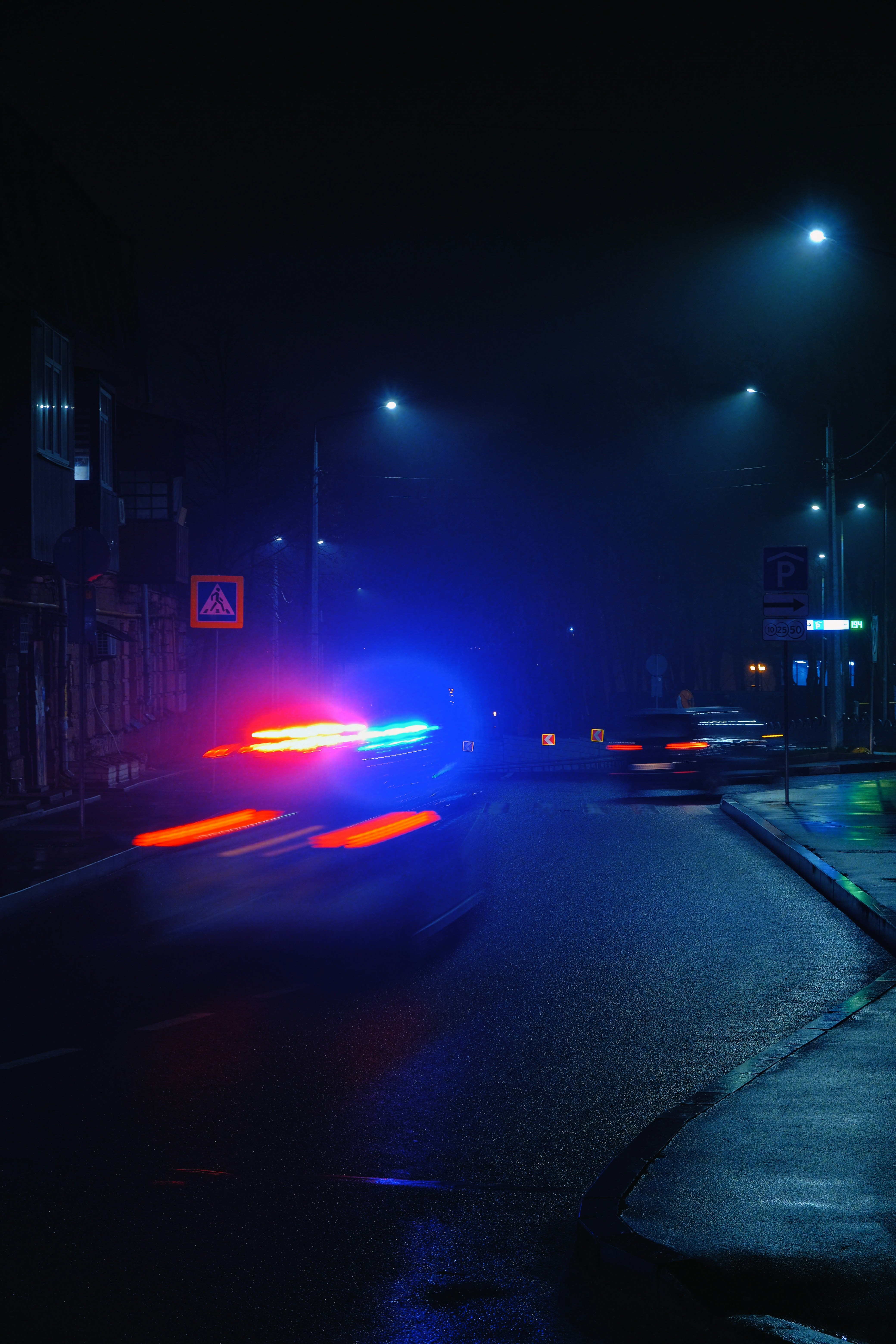 Police car with flashing lights speeding down a wet street at night, illuminated by street lamps and reflecting off the pavement.