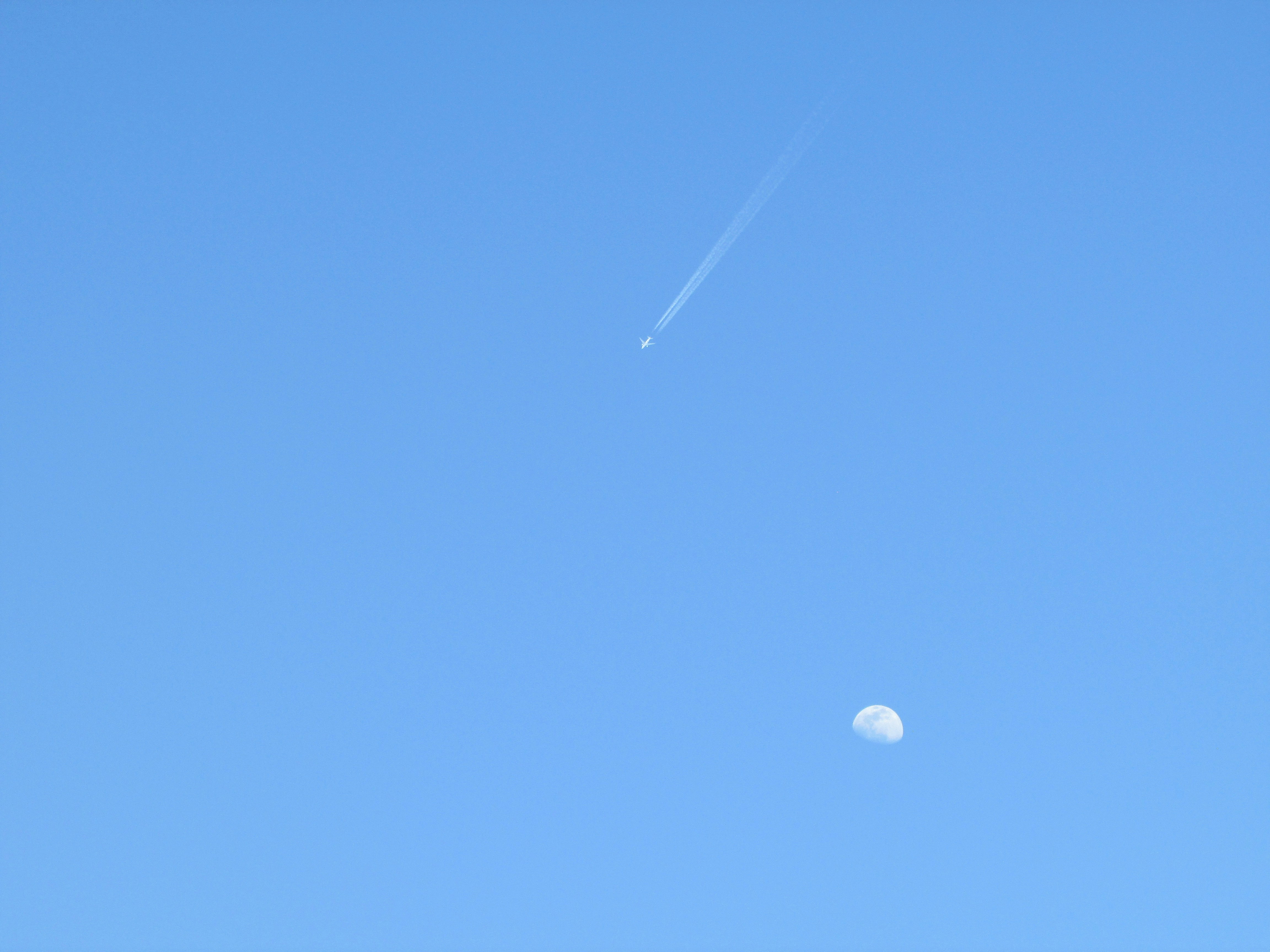 A jet trails through a clear blue sky, while the moon hangs serenely below, creating a juxtaposition of human flight and celestial presence.