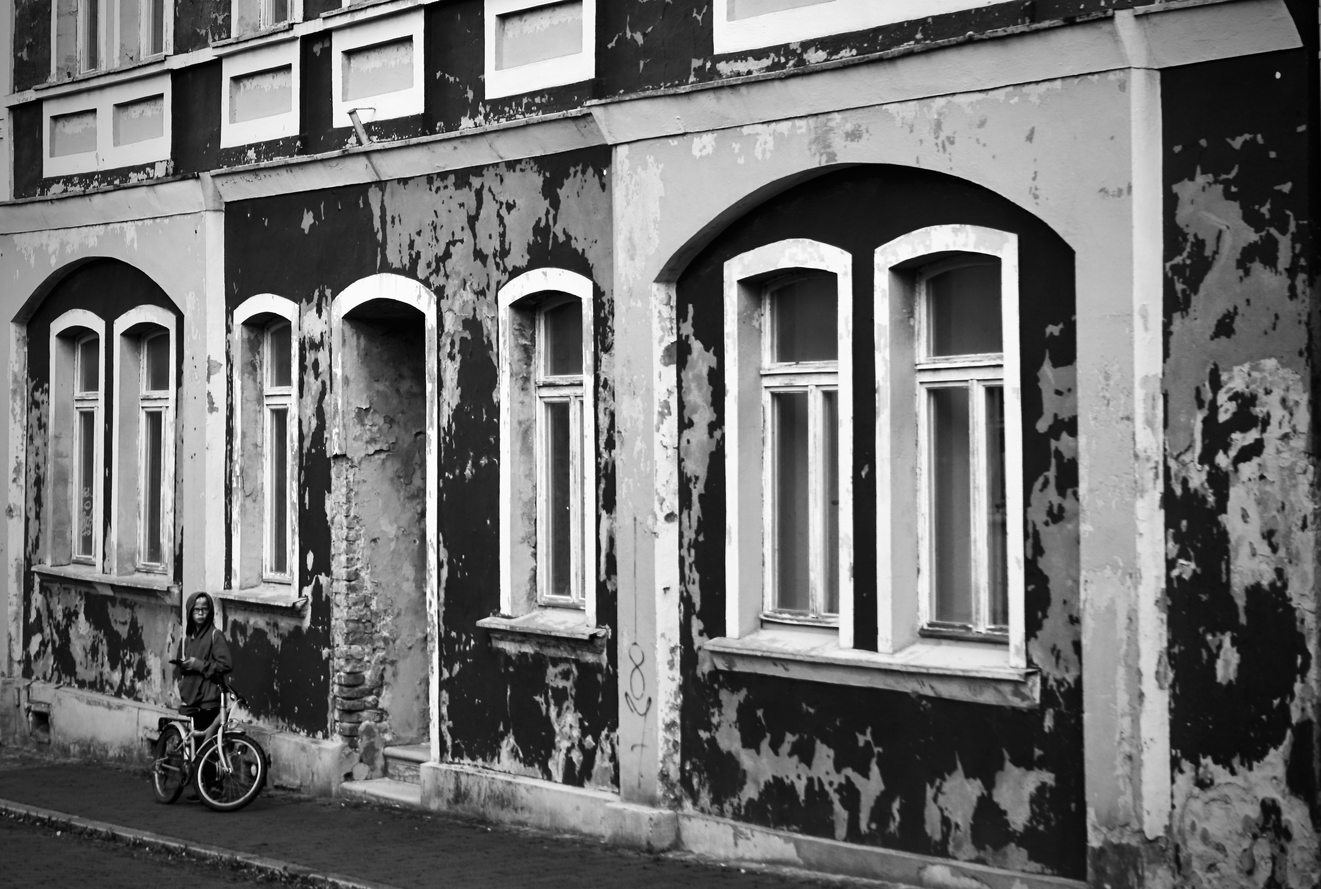 A young child on a bicycle stands beside a weathered, peeling wall adorned with multiple windows. The monochrome tones enhance the nostalgic atmosphere.