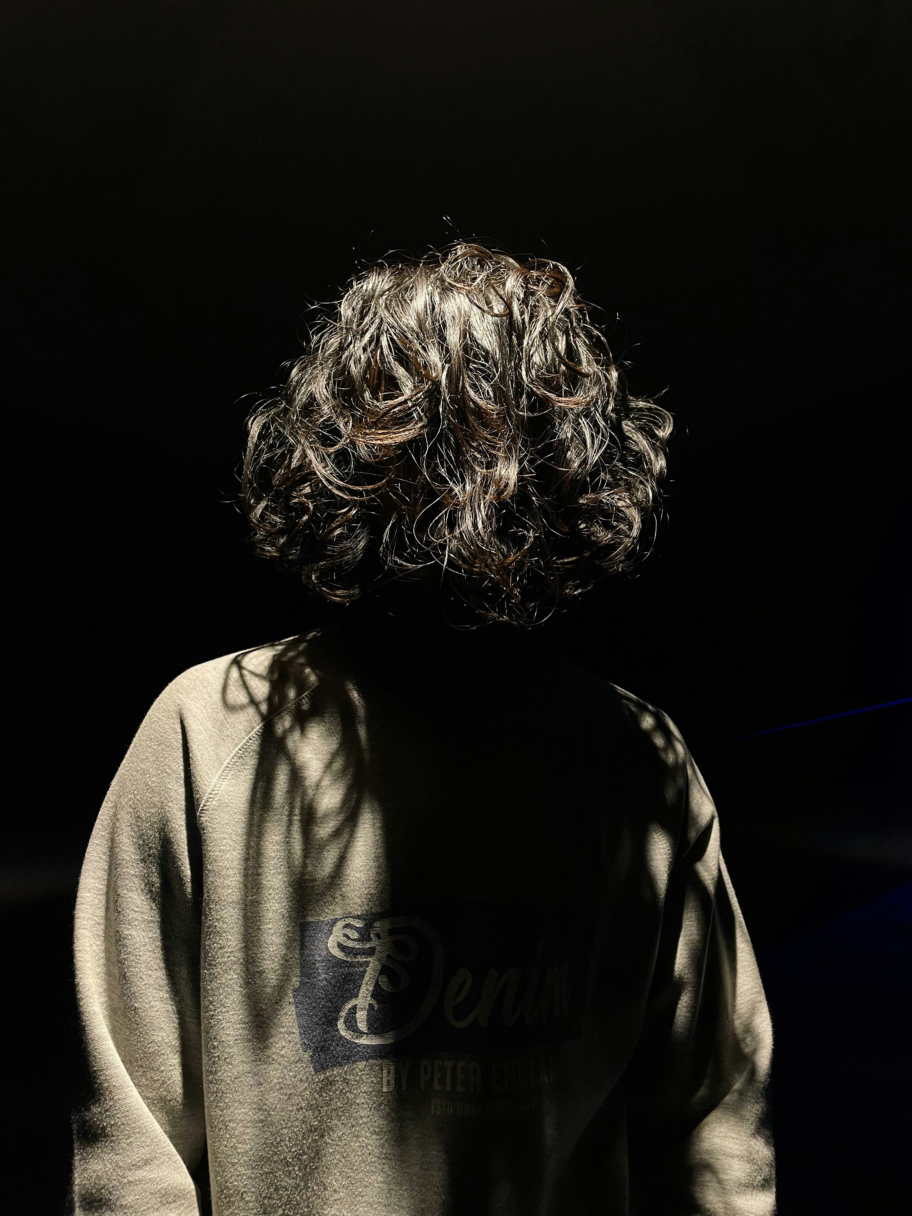 Curly-haired figure stands in shadow, illuminated by a subtle light source, creating a dramatic silhouette effect.