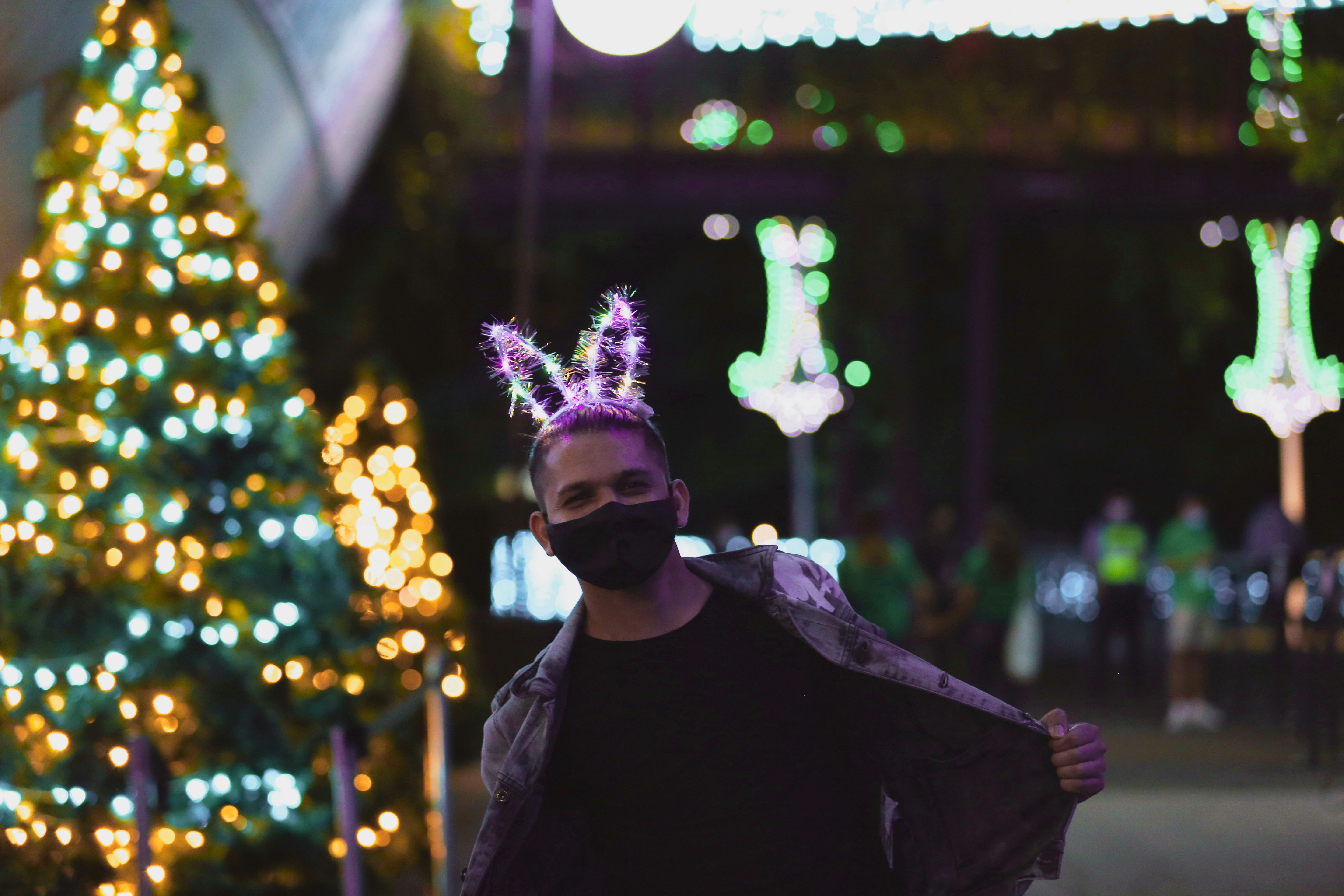 A young man wearing a mask and decorative bunny ears poses playfully in a festive setting adorned with colorful lights and a Christmas tree.