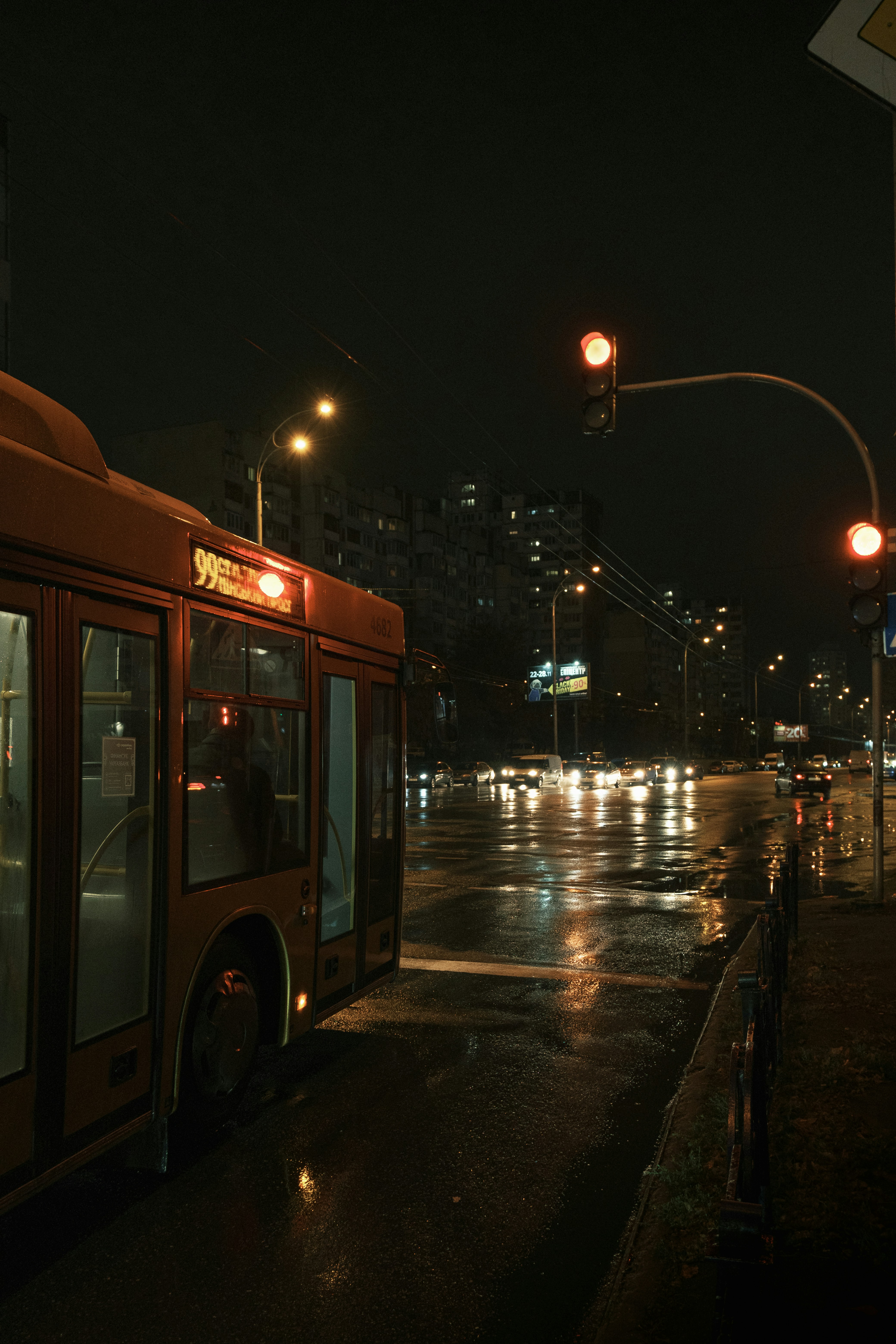 An orange bus waits at a traffic light on a rainy night, illuminated by streetlights and the glow of city traffic. The wet pavement reflects the vibrant lights.