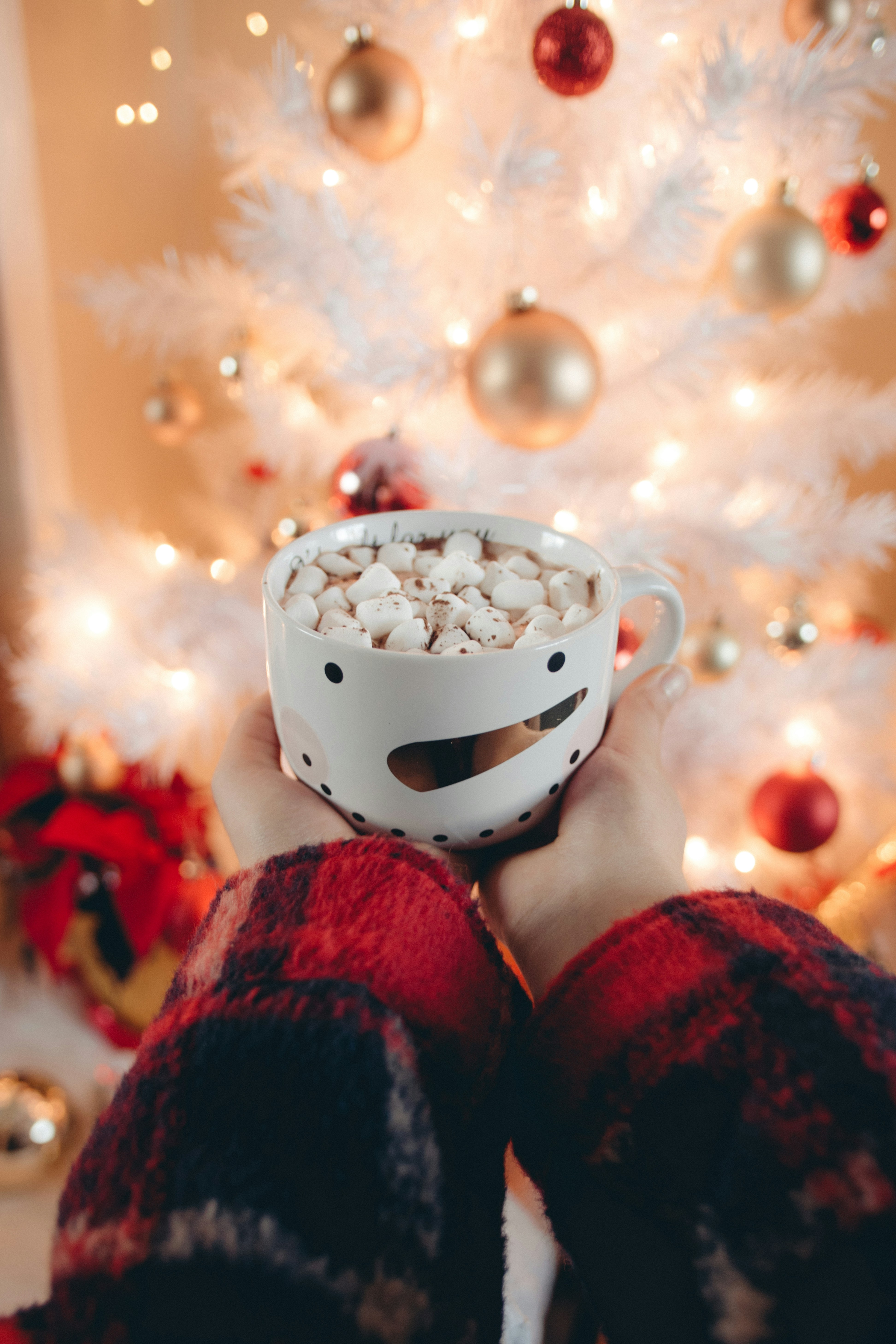 Hands holding a cheerful mug filled with hot chocolate and marshmallows, set against a festive white Christmas tree adorned with ornaments.