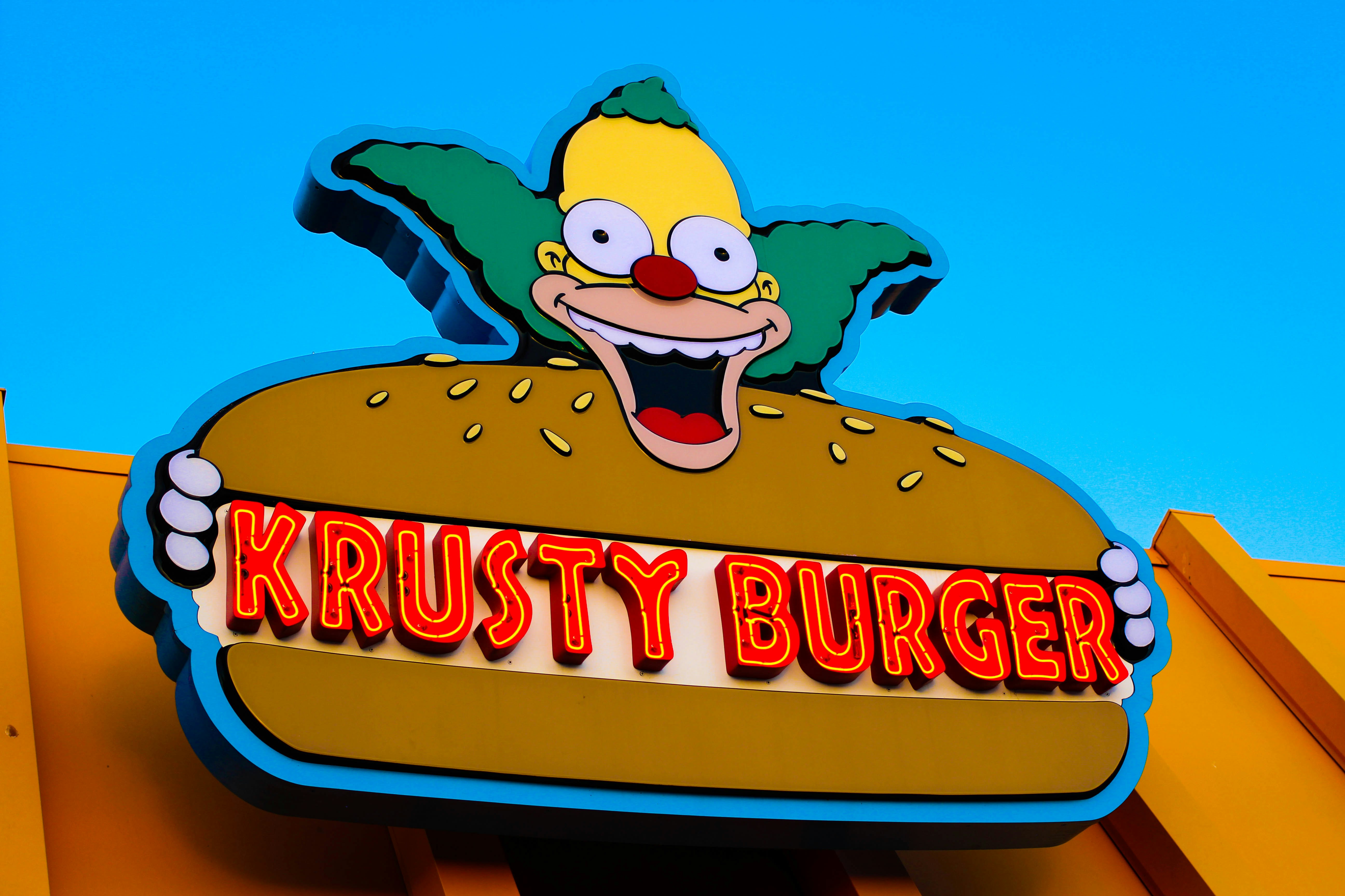 Brightly colored Krusty Burger sign featuring a cartoon character with a clownish grin, set against a clear blue sky.