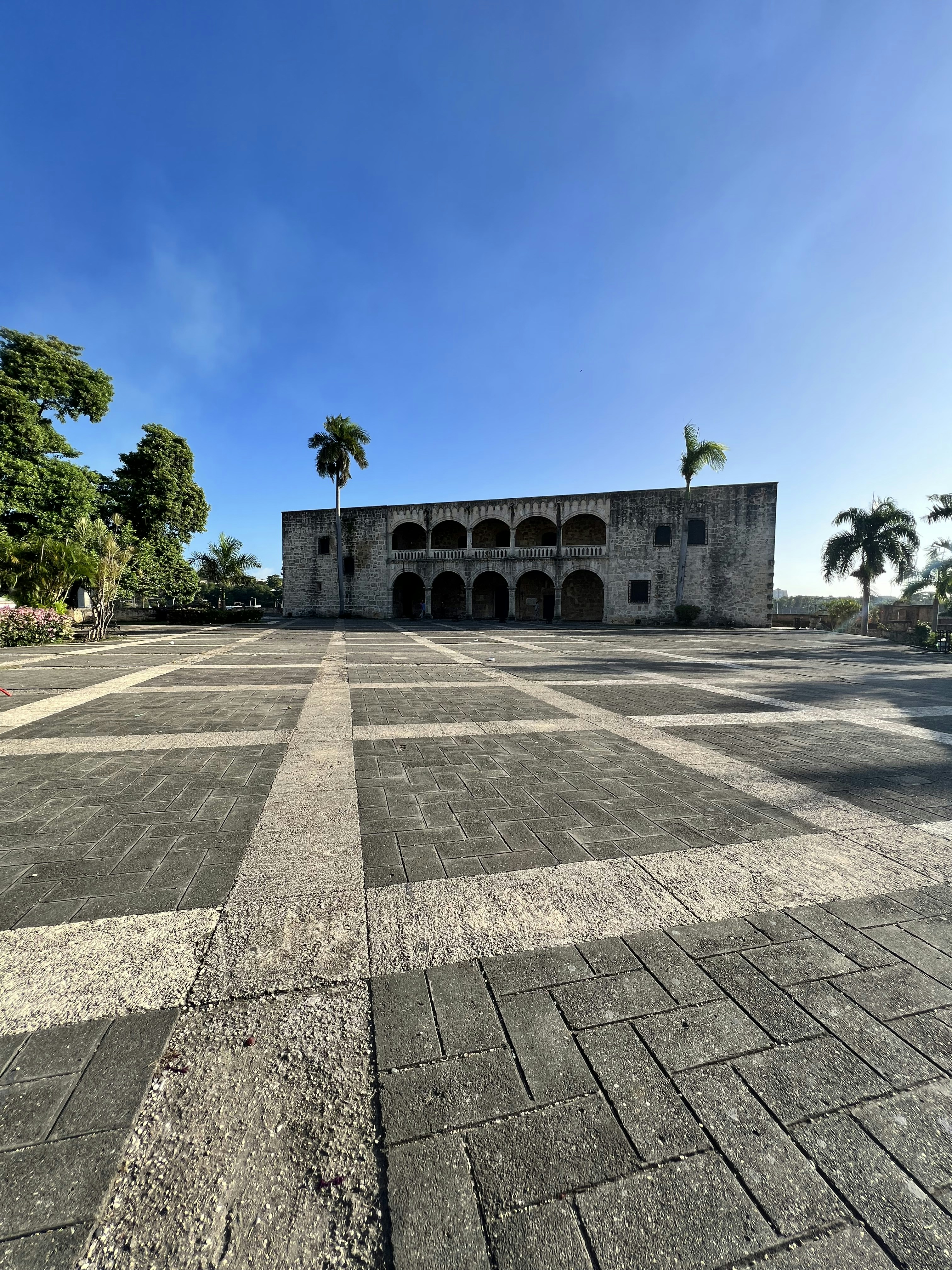Historic stone building with arched windows, surrounded by palm trees and a patterned stone courtyard under a clear blue sky.