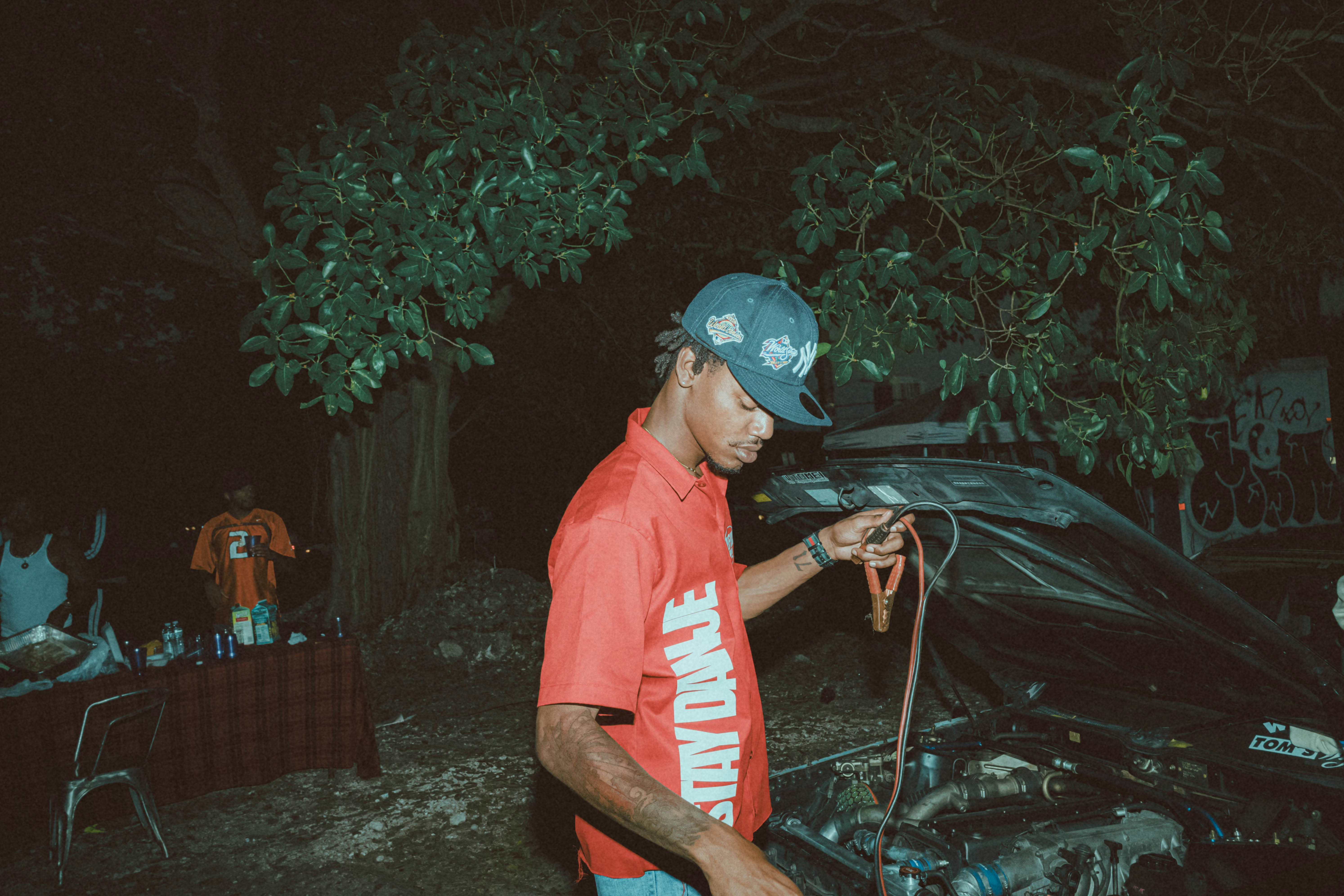Young mechanic working on a car engine under the night sky, surrounded by friends and a lively atmosphere.