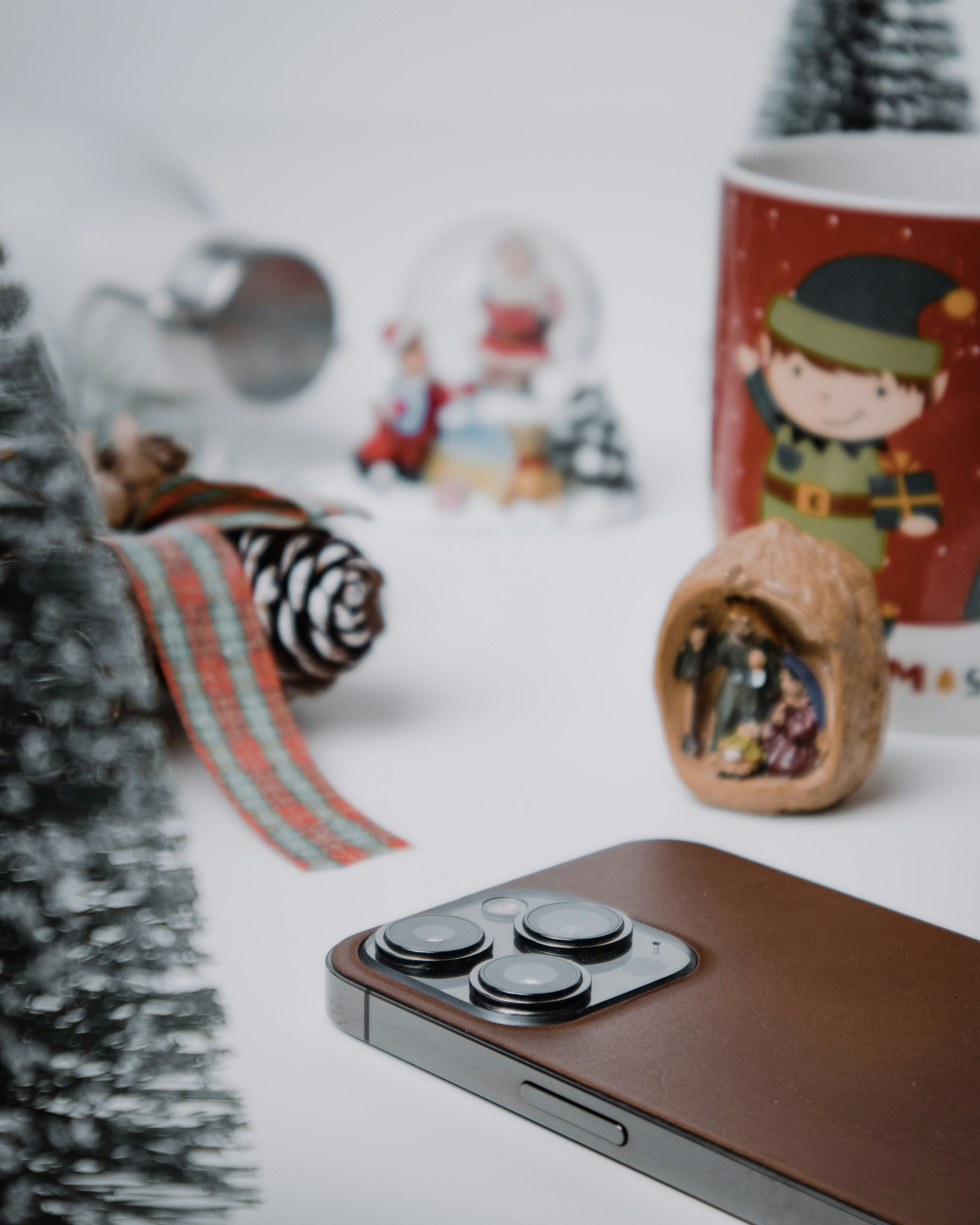 A smartphone rests among holiday decorations, including a festive mug, a snow globe, and pinecones, creating a cozy seasonal atmosphere.