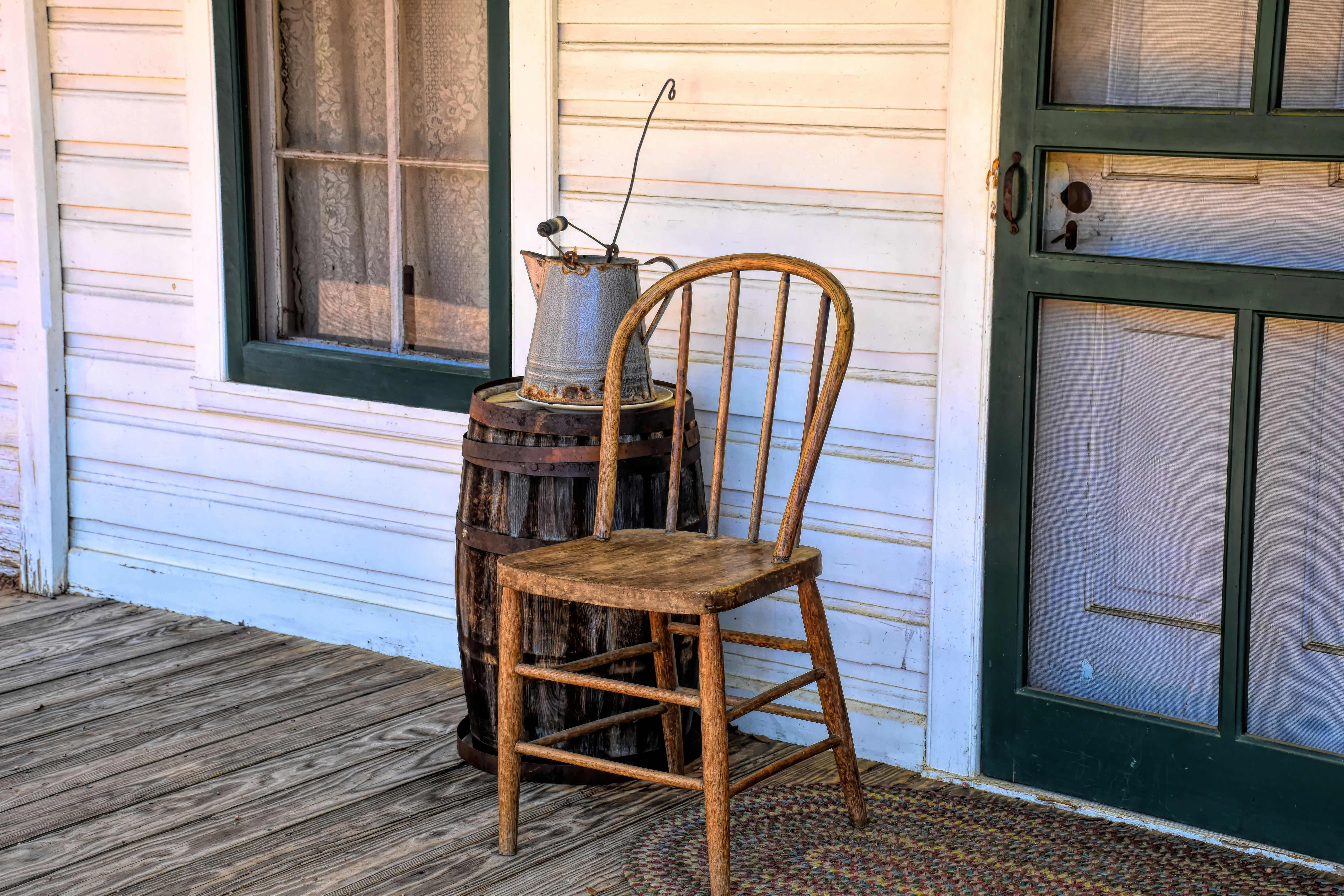 Wooden chair beside a weathered barrel and a vintage watering can on a porch. The scene evokes nostalgia and a sense of tranquility.