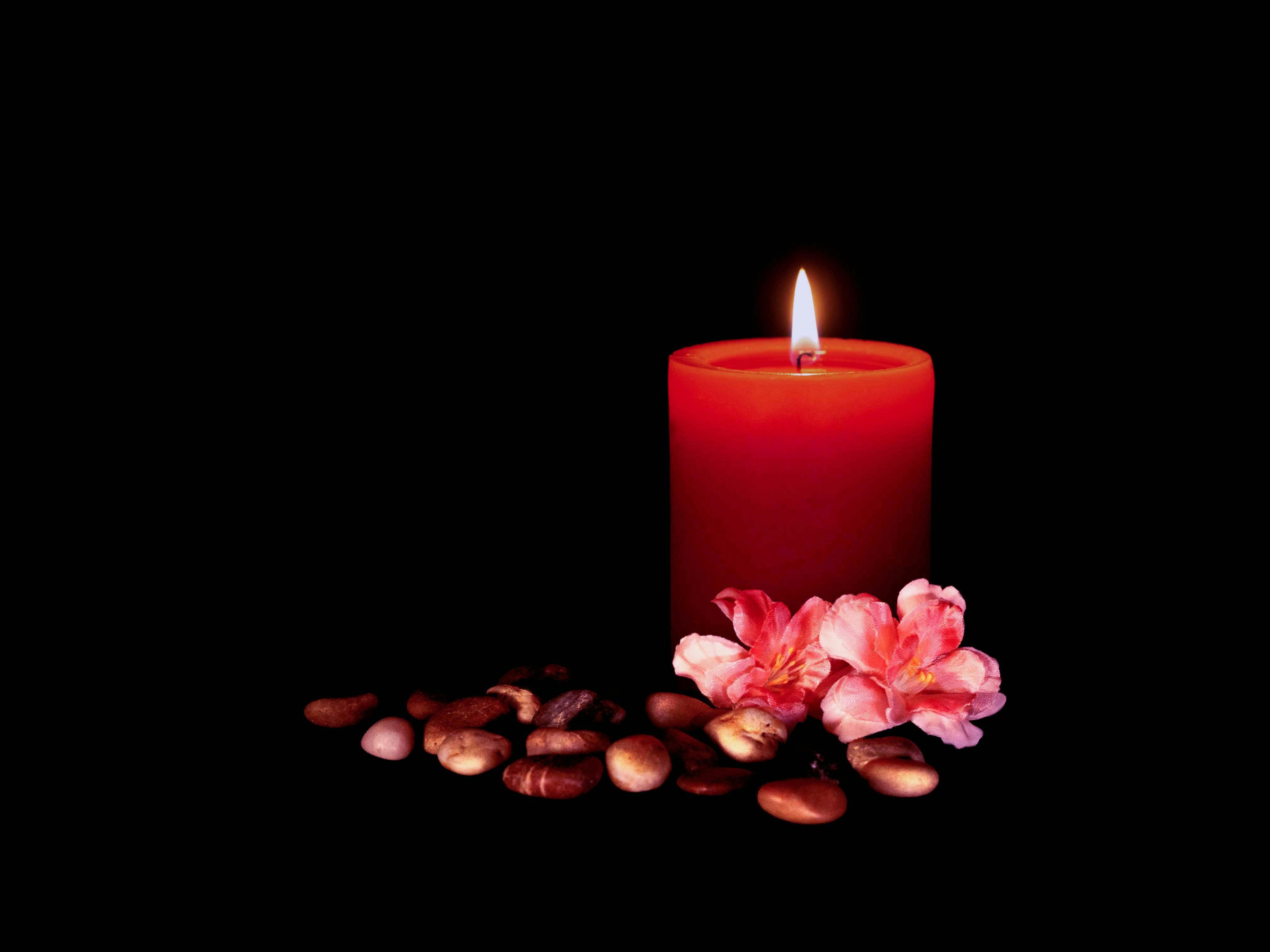 A red candle emits a warm glow, surrounded by smooth pebbles and delicate pink flowers against a black backdrop.