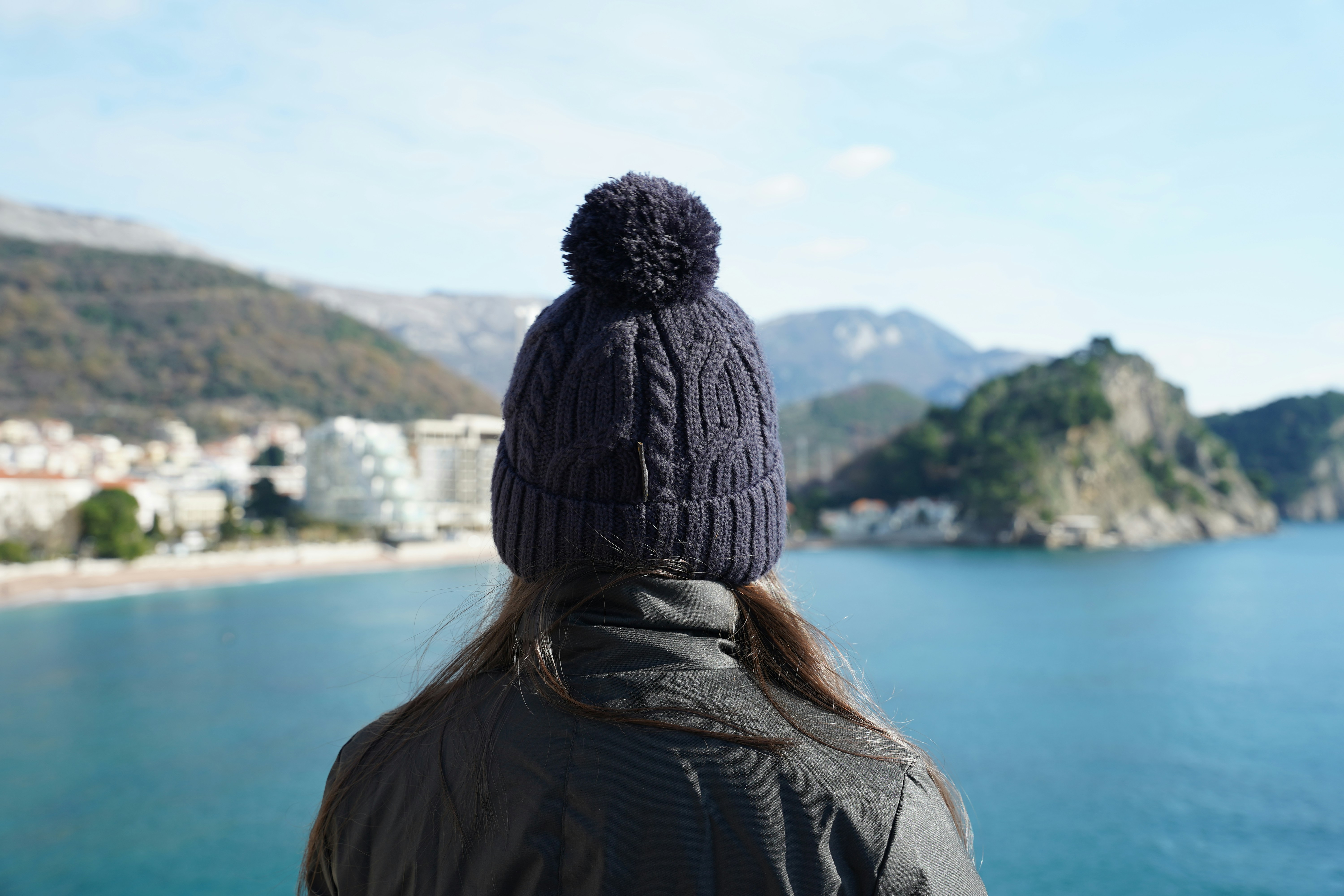 A person in a knitted hat gazes out over a tranquil coastal landscape, with mountains and a distant island framing the view.