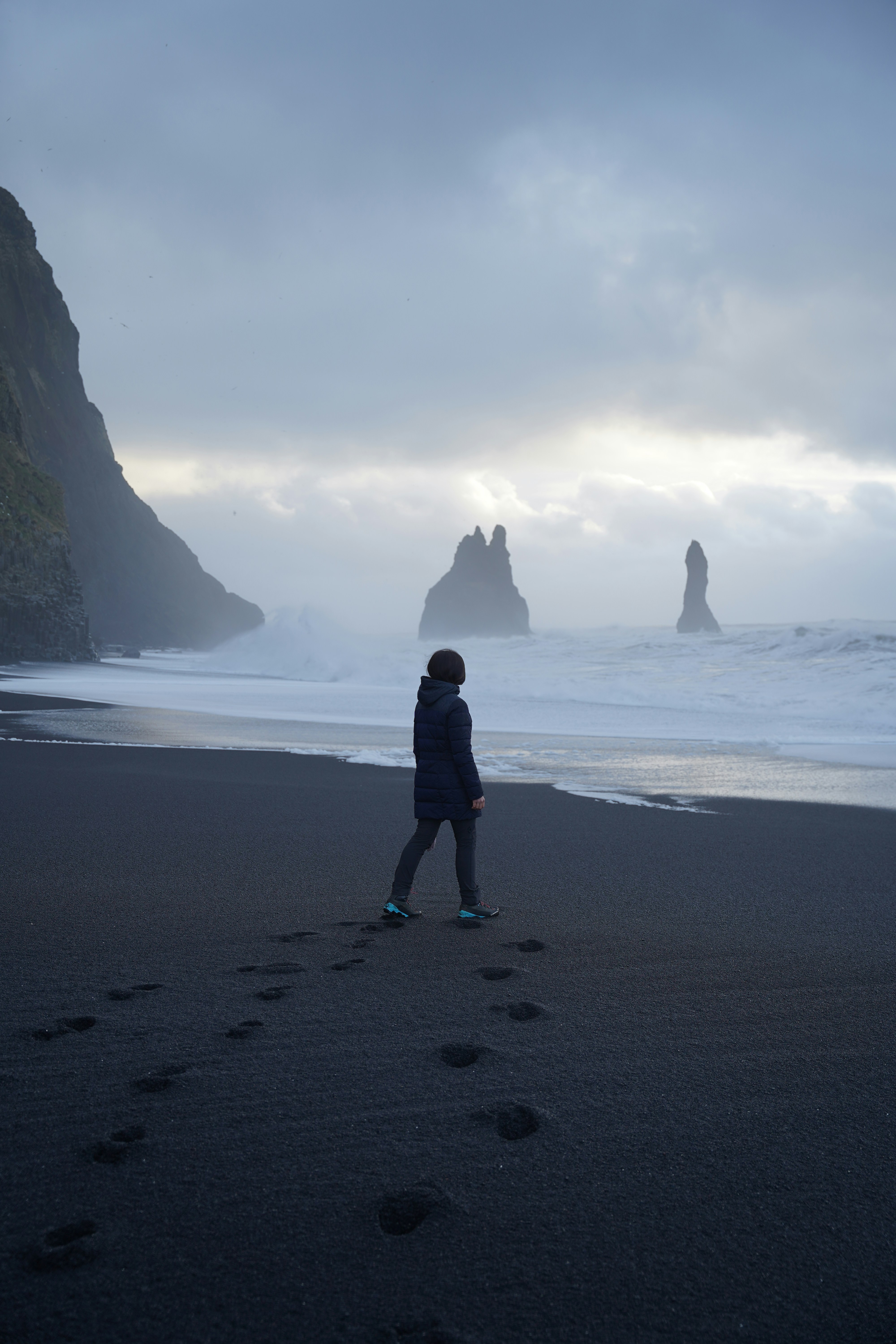 Person walking along a black sand beach with towering sea stacks in the distance under a moody sky.