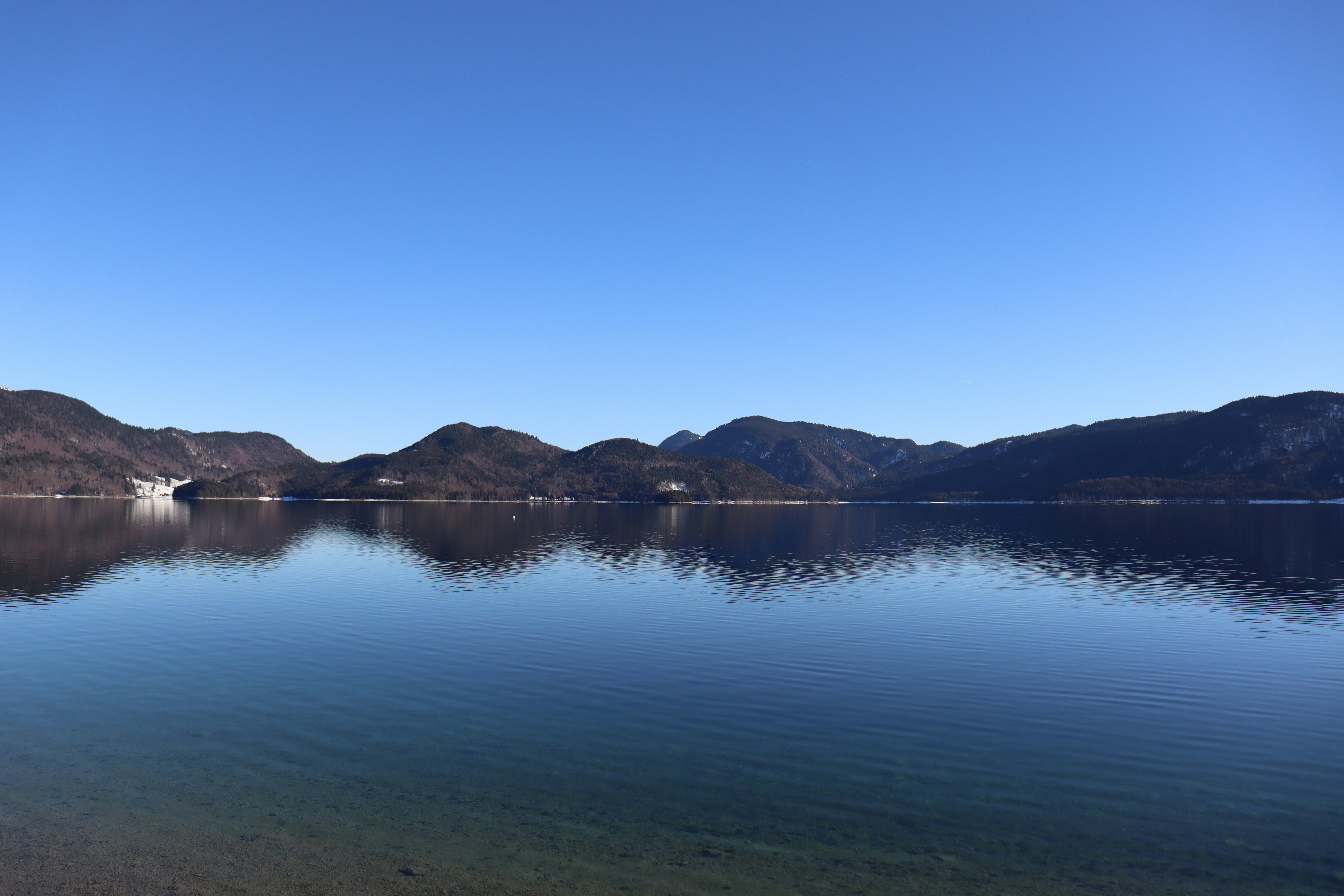 Calm lake reflecting distant mountains under a clear blue sky. The tranquil scene invites contemplation.