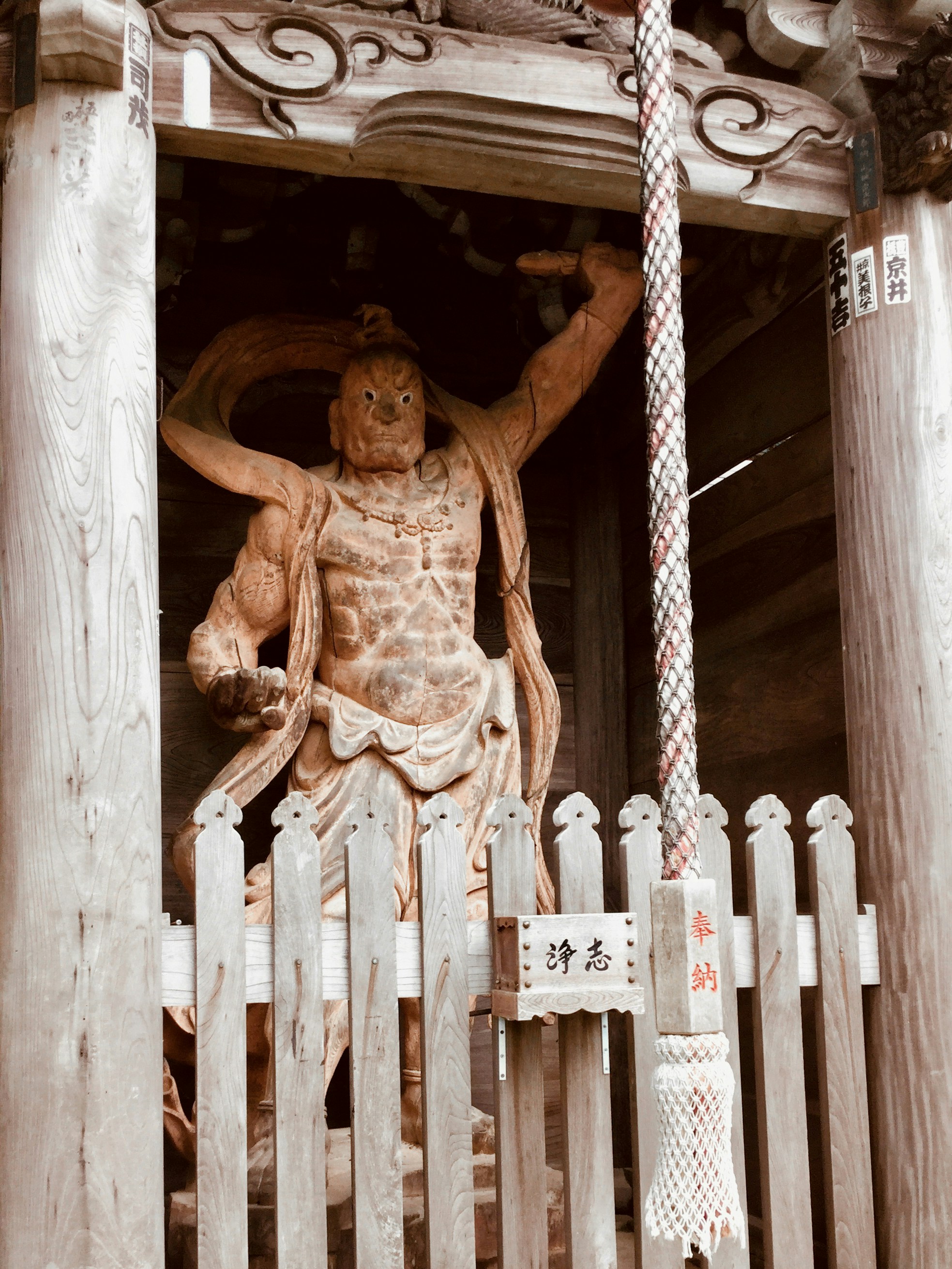 Intricately carved wooden statue of a guardian deity, poised within a wooden shrine, showcasing detailed craftsmanship. The figure exudes a sense of strength and protection.