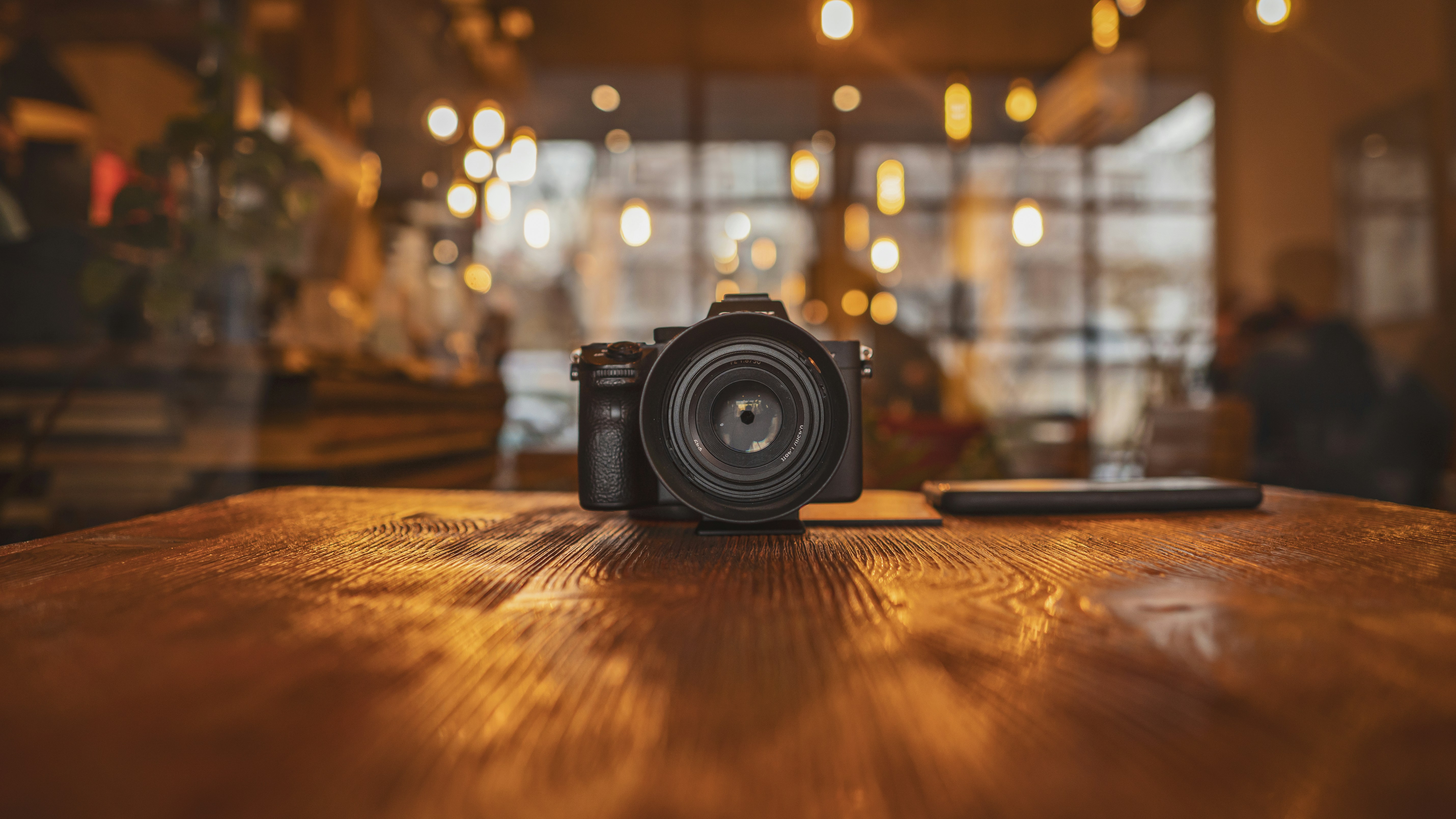 A sleek camera rests on a wooden table, surrounded by a cozy café ambiance with warm lighting and blurred patrons in the background.