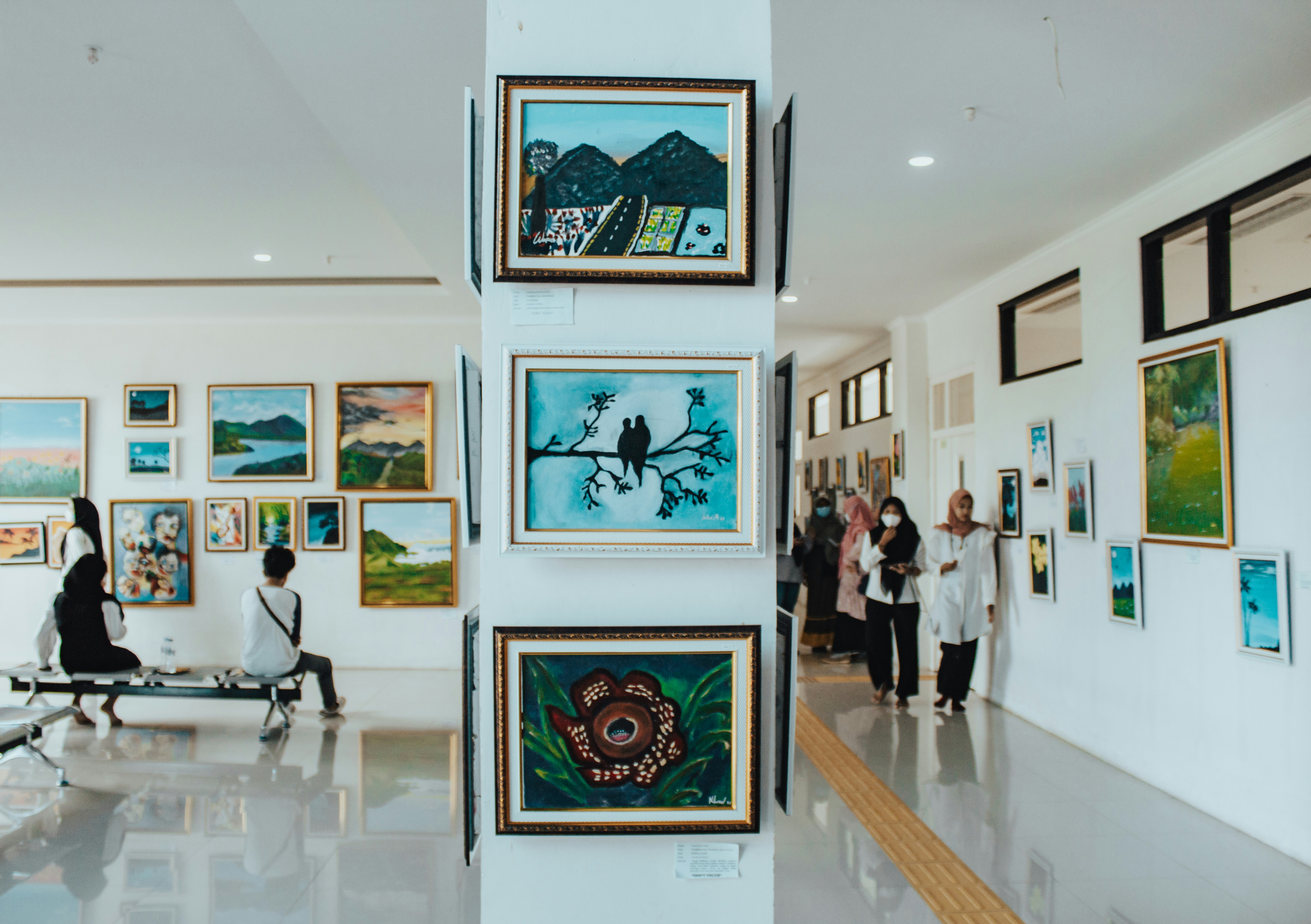 Art gallery with various paintings displayed on walls and a central pillar, visitors browsing the exhibits.