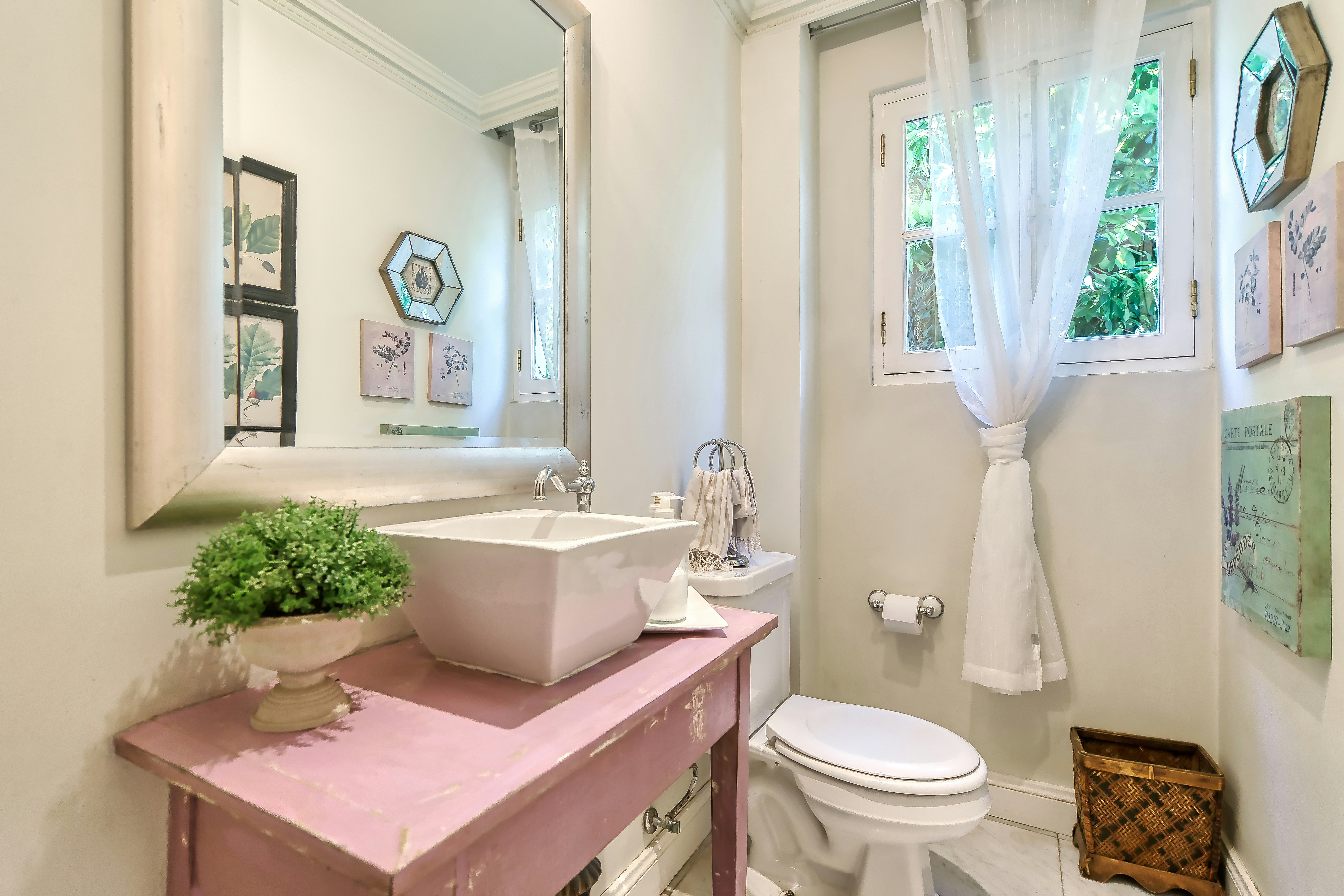 Bright bathroom featuring a stylish vessel sink on a pink vanity, framed by soft curtains and decorative wall art.