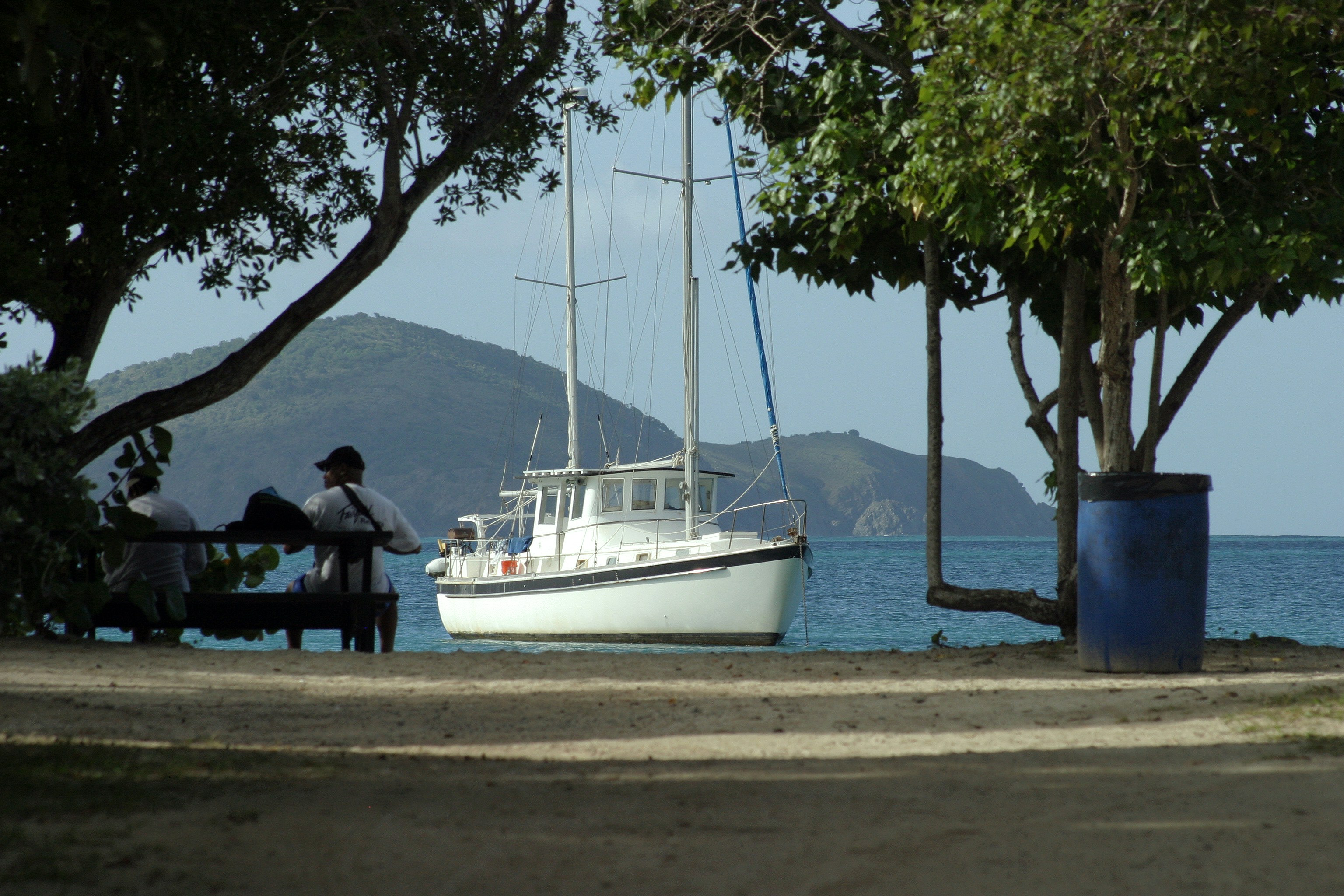 A sailboat anchored near a tranquil beach, framed by lush trees and distant hills. The scene captures a moment of relaxation and connection with nature.