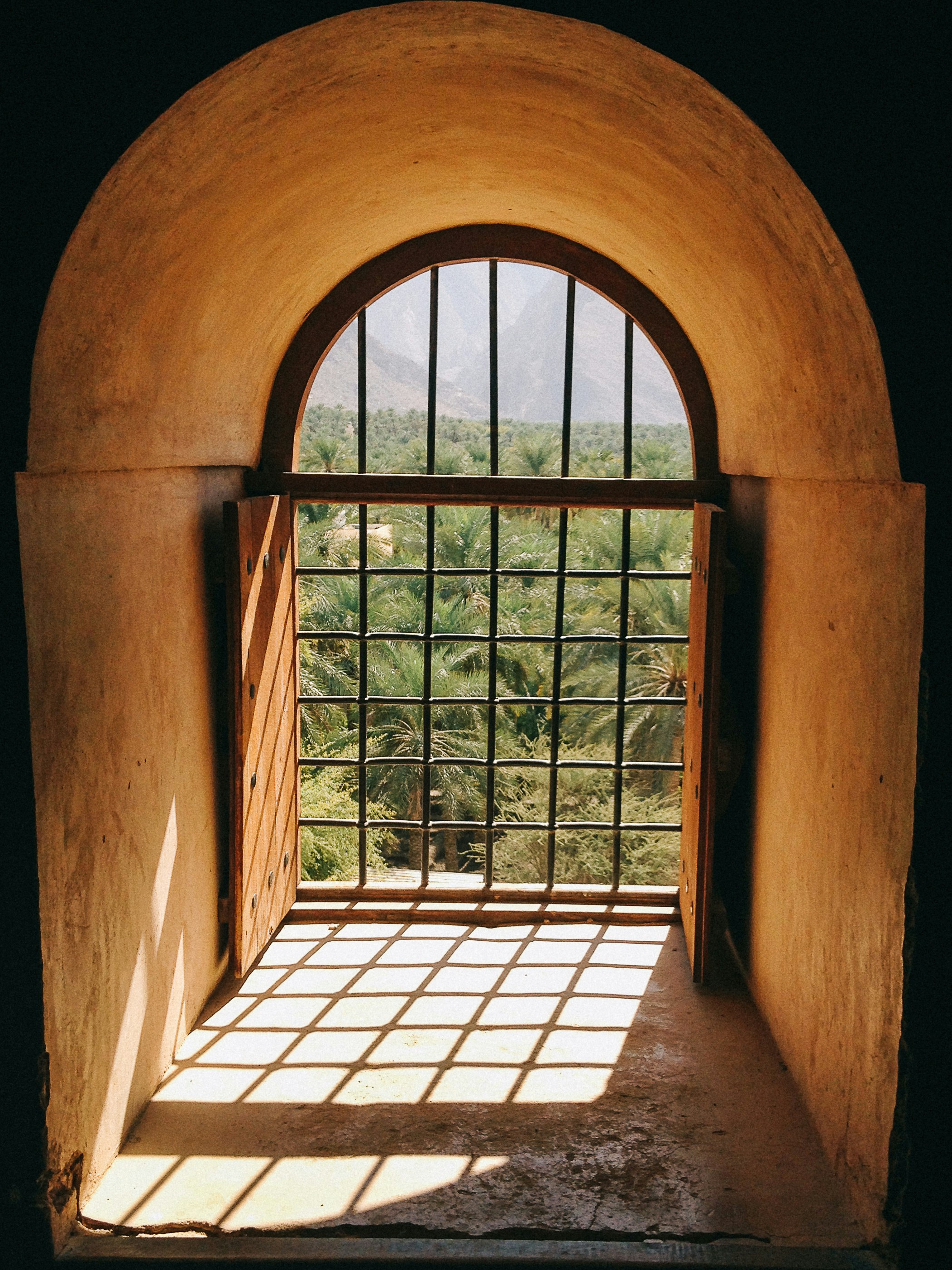 Photograph of an arched window with a metal grid opening to a verdant valley. Warm sunlight casts striped shadows across the stone floor.