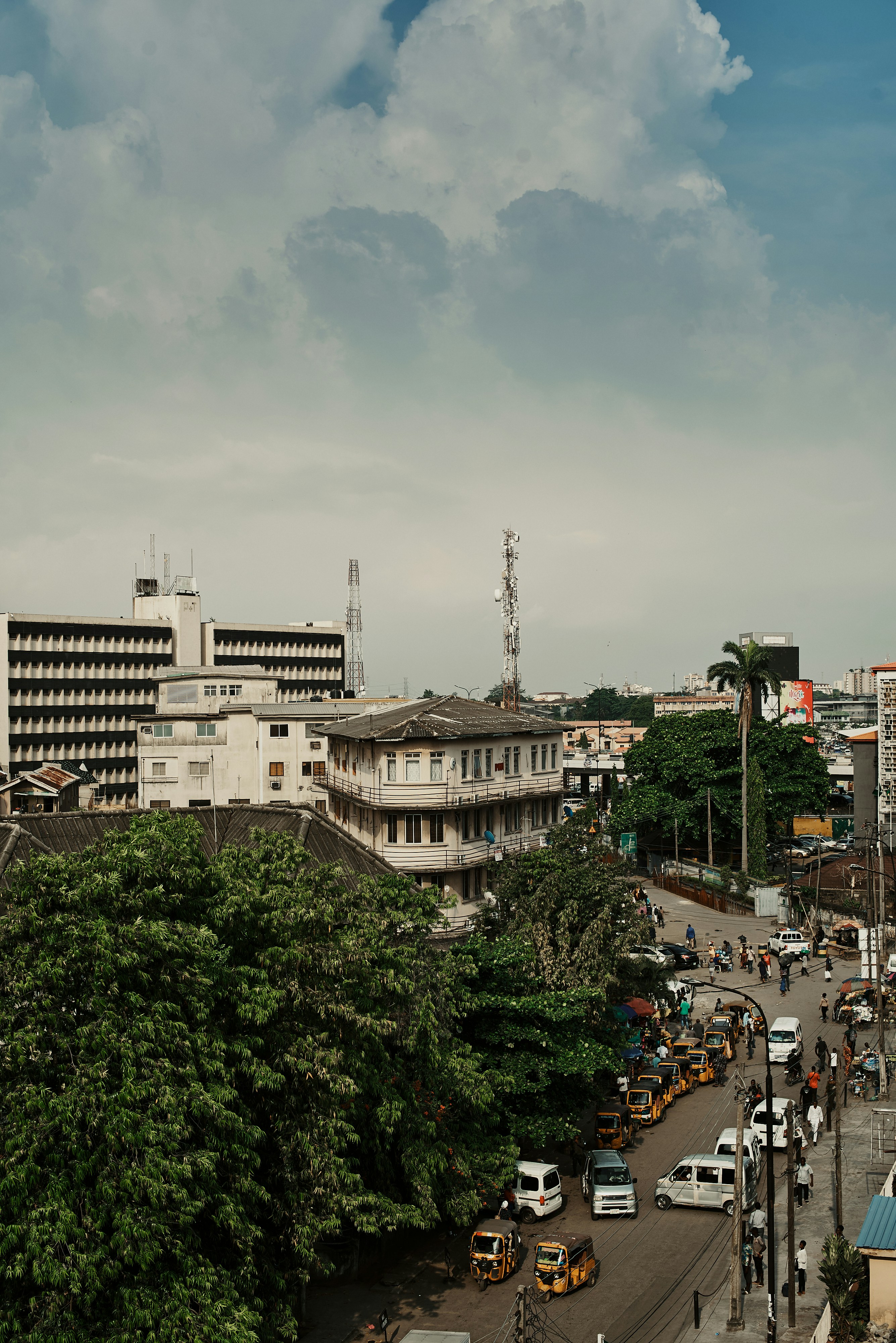 A busy city intersection with vibrant green trees and a mix of vehicles, showcasing the dynamic urban landscape. The scene captures the essence of daily life in a metropolitan area.