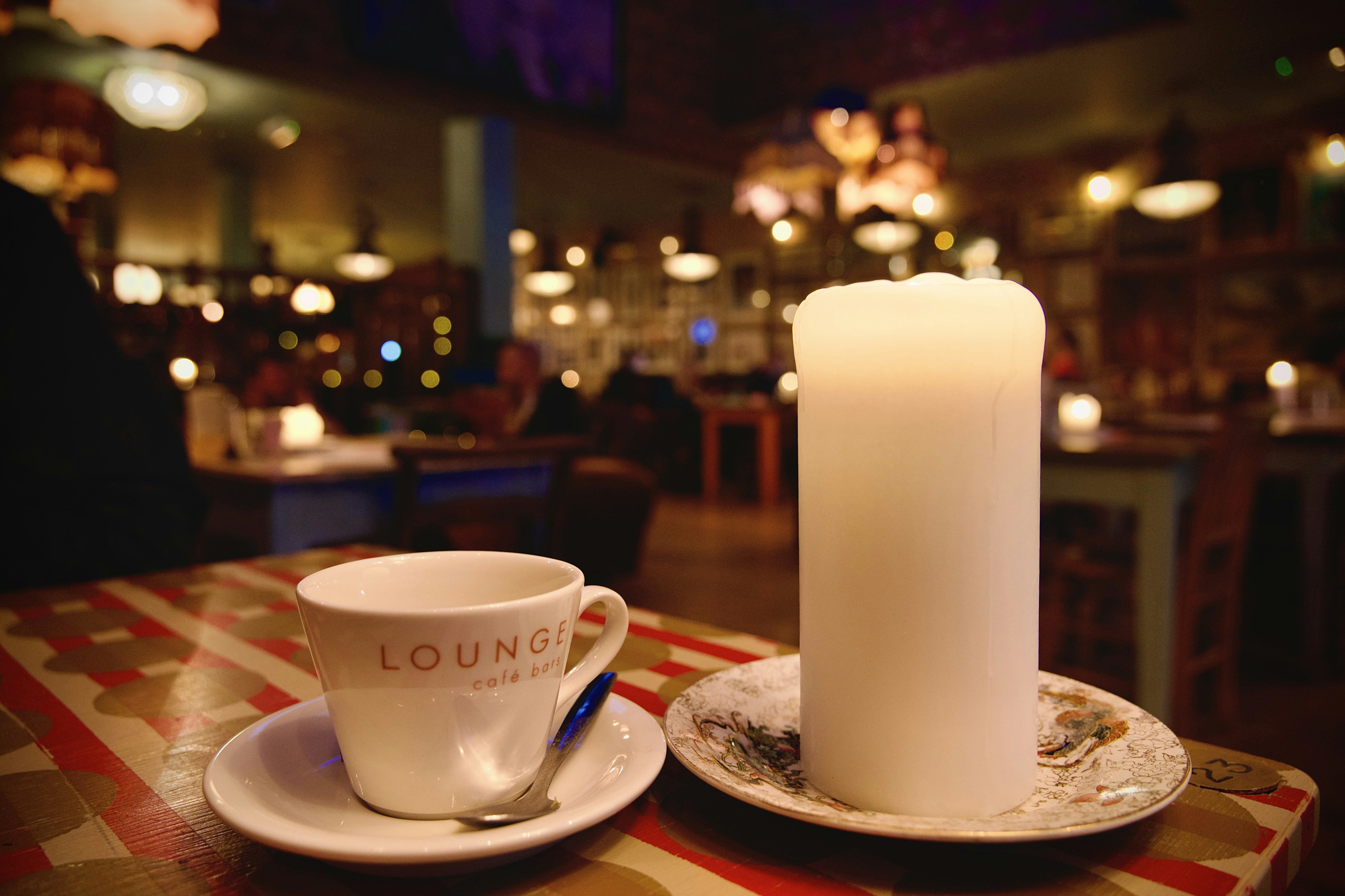 A white candle and a coffee cup sit on a patterned table in a warm café atmosphere, inviting relaxation and conversation.
