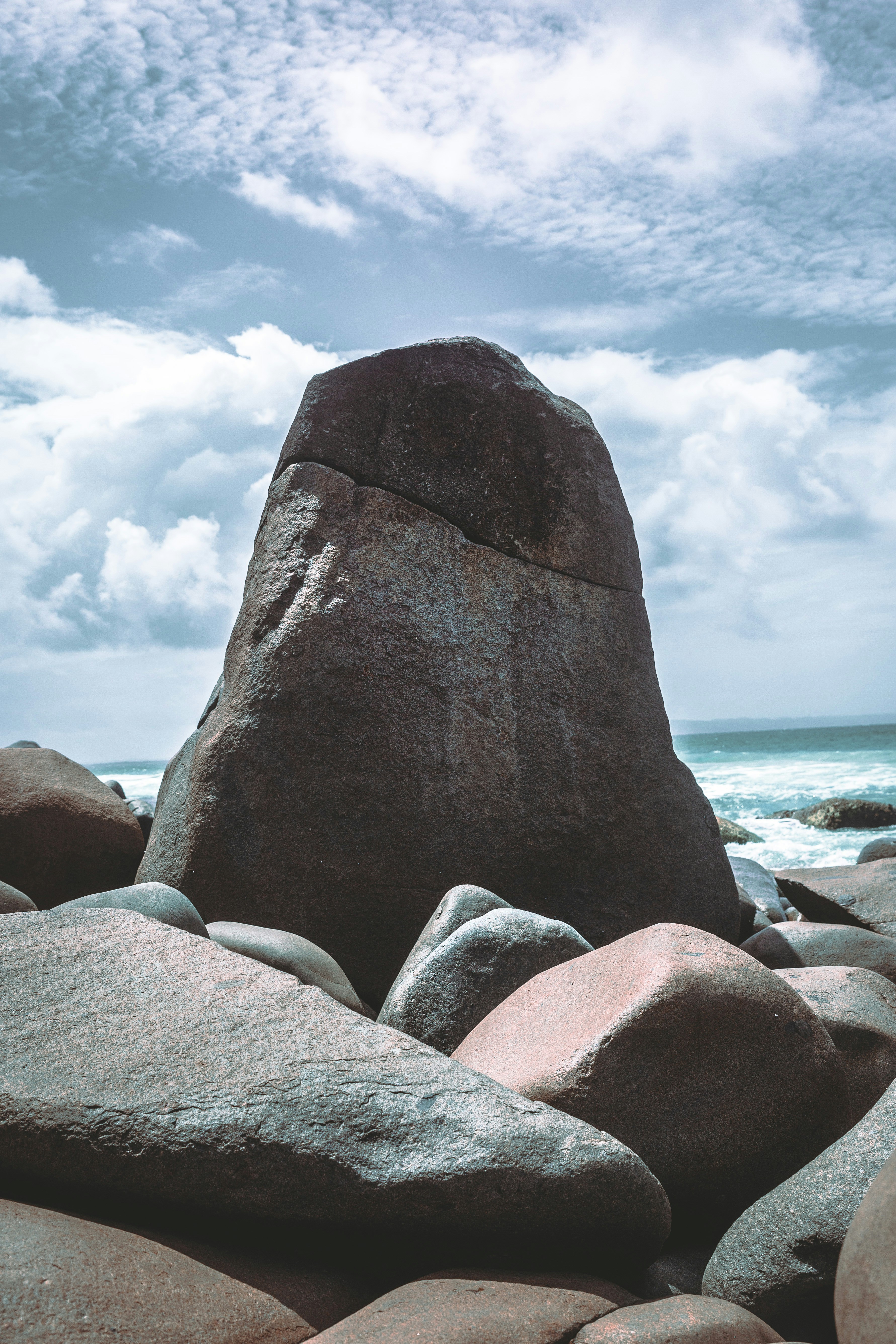 A towering rock formation stands resilient among a scatter of smooth stones, framed by a dramatic sky and crashing waves.