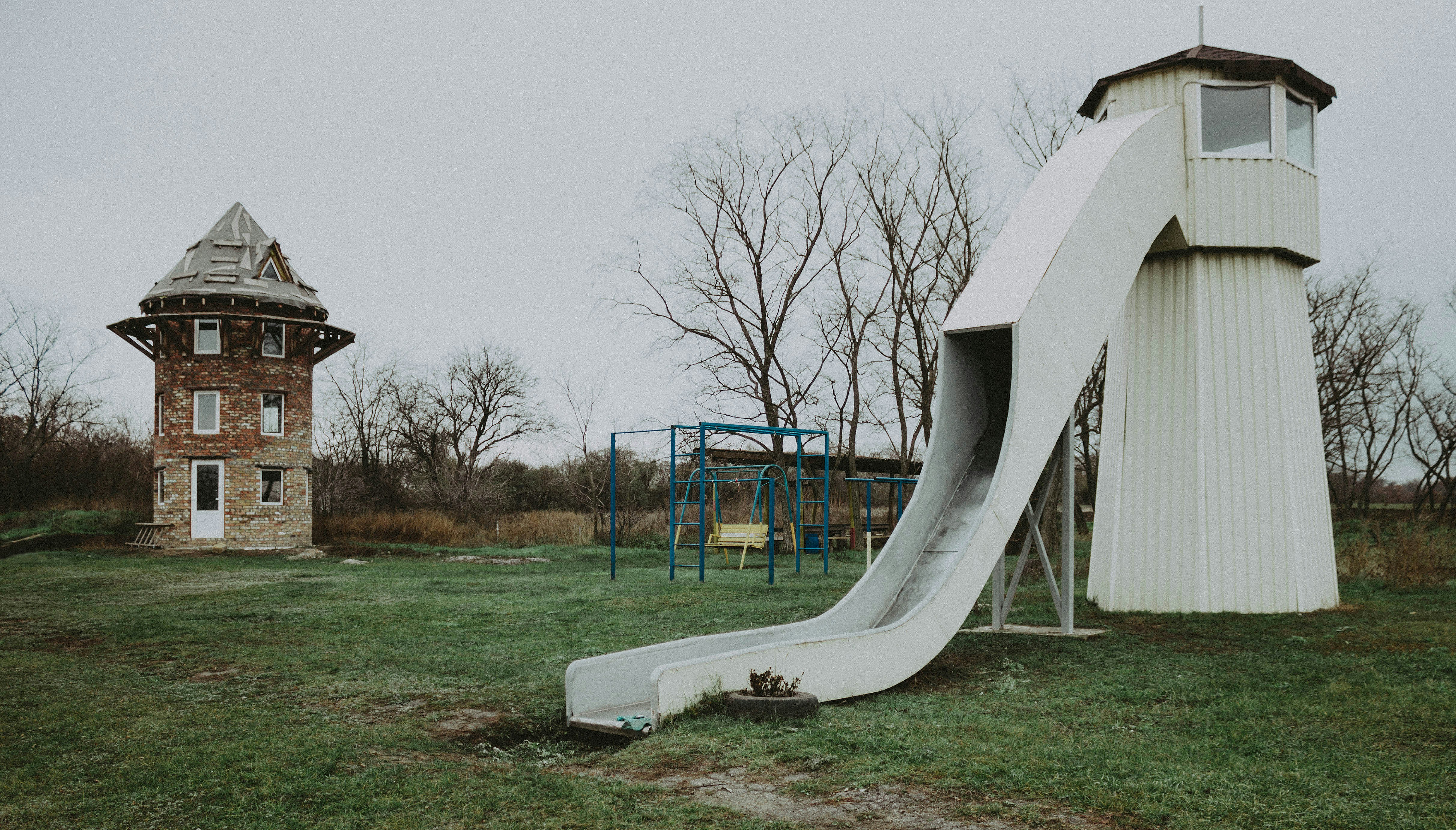 An unconventional playground featuring a unique slide and a tower resembling a whimsical building, surrounded by a grassy area and sparse trees.