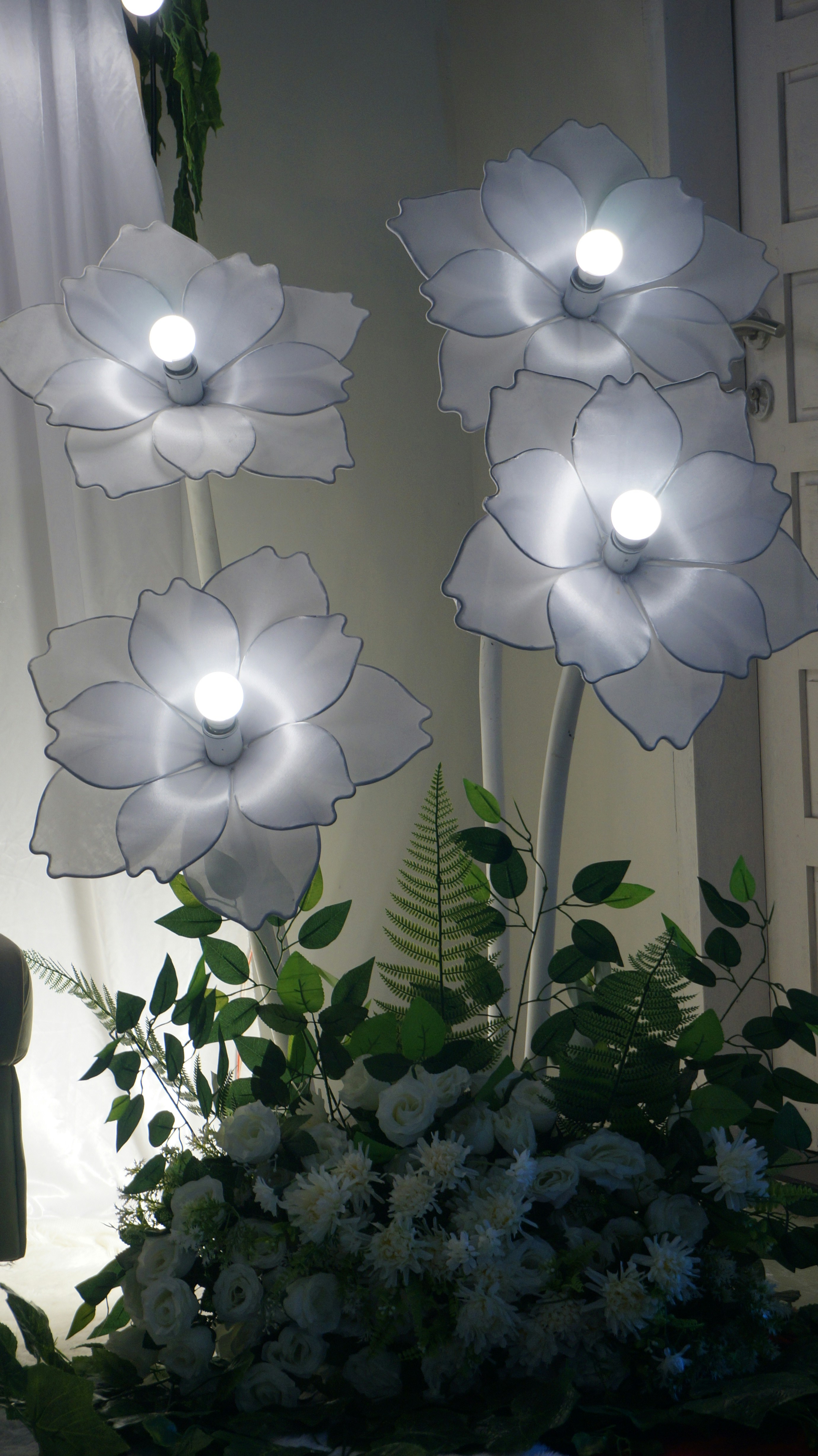 White flower-shaped lamps on tall stems illuminate a lush indoor arrangement of greenery and white blooms.