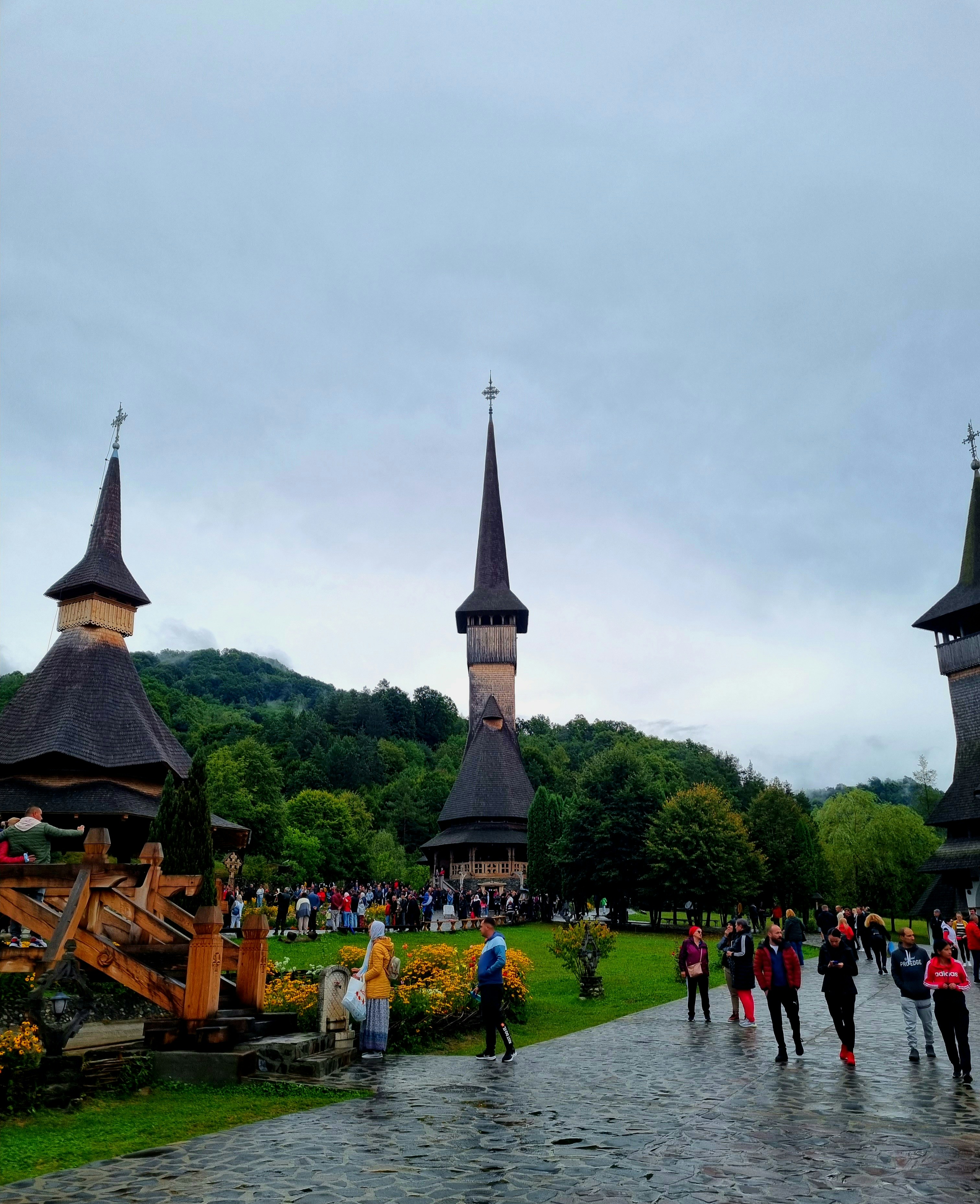 Visitors explore a scenic park featuring traditional wooden churches with tall spires, surrounded by lush greenery and vibrant flowers.
