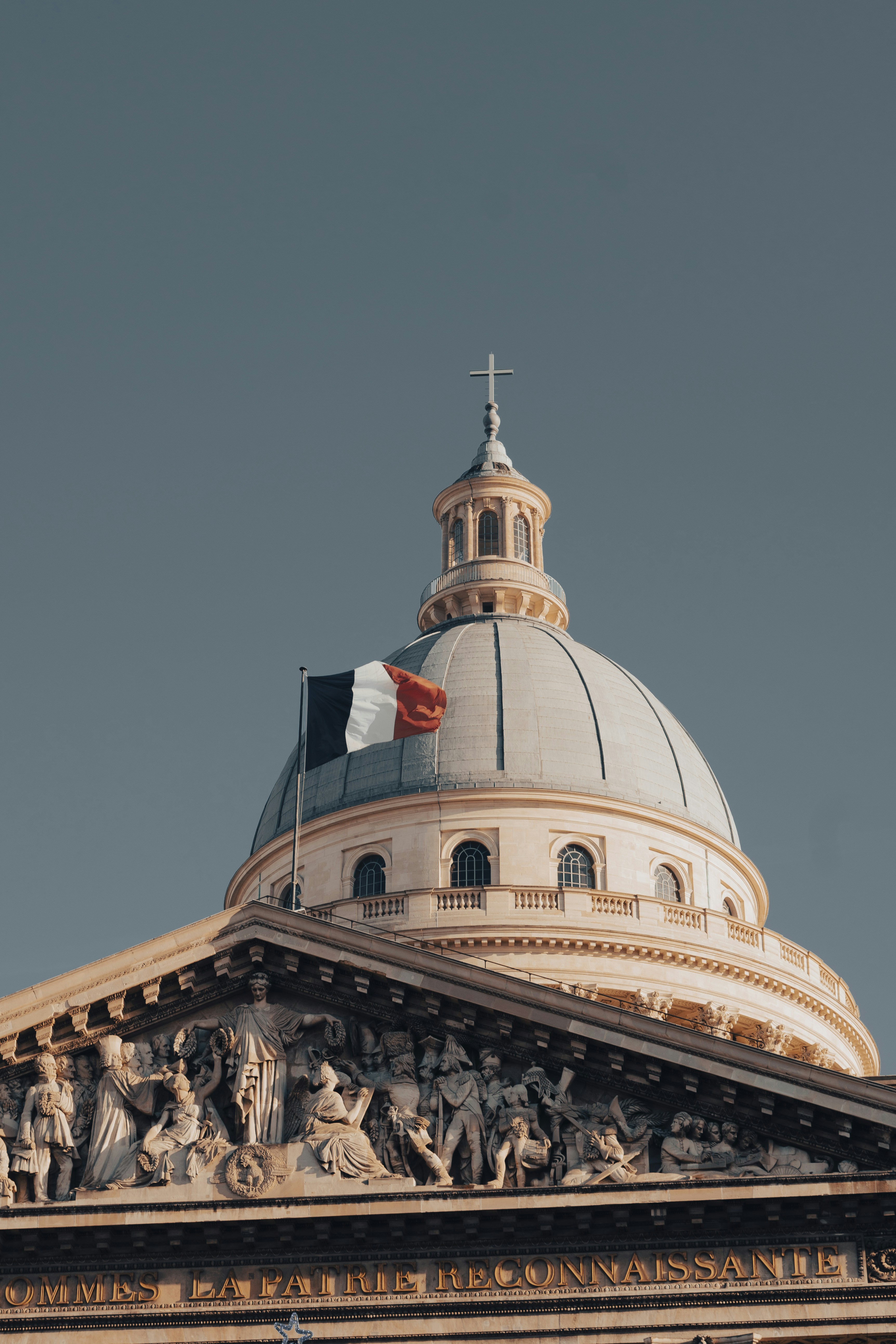 The ornate dome of a historic building, crowned with a cross and flanked by a waving French flag. Intricate sculptures adorn the facade, reflecting cultural significance.