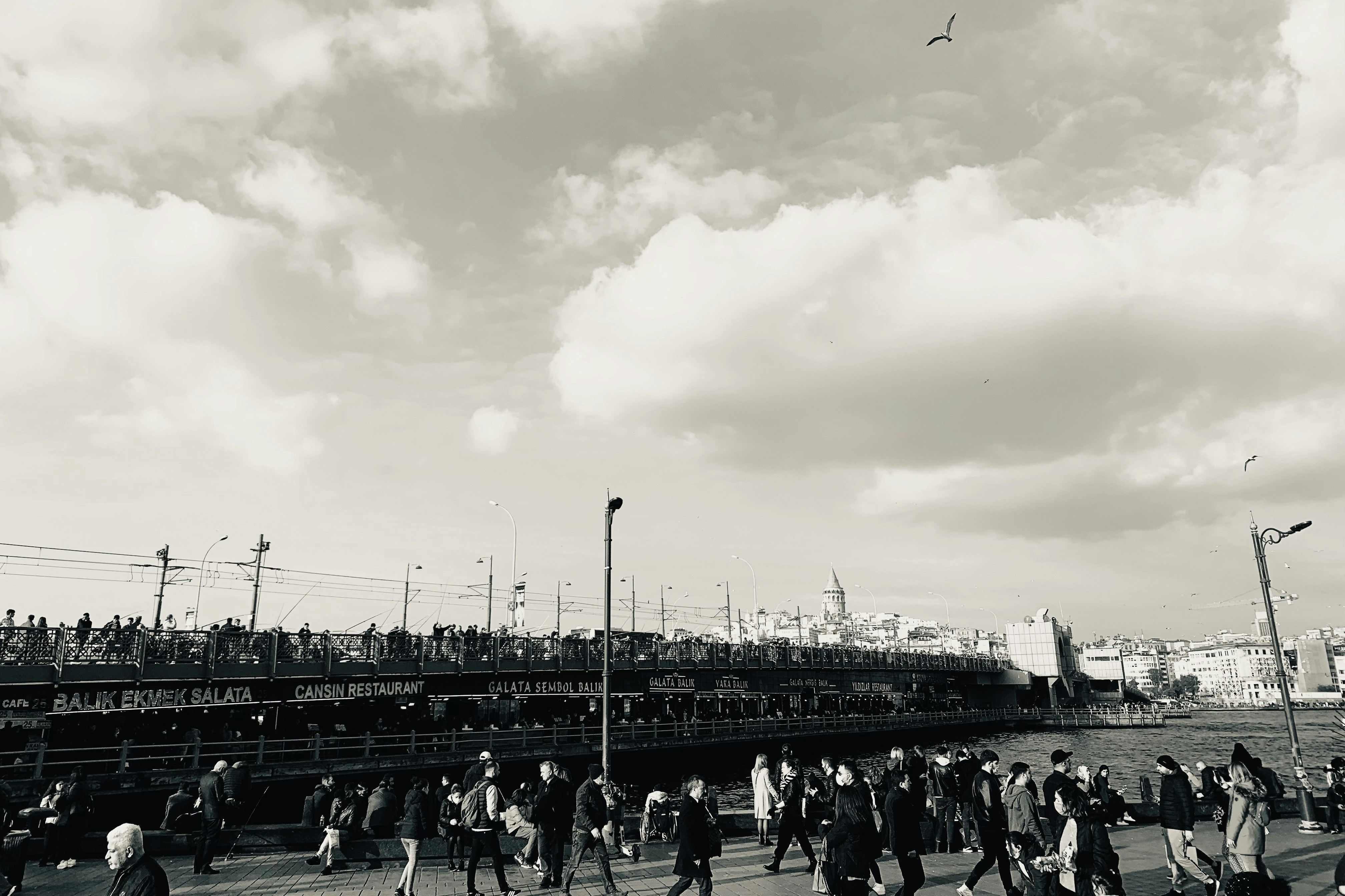 Crowd of pedestrians bustling along a waterfront promenade with a busy pier in the background. The scene is set against a dramatic sky filled with clouds.