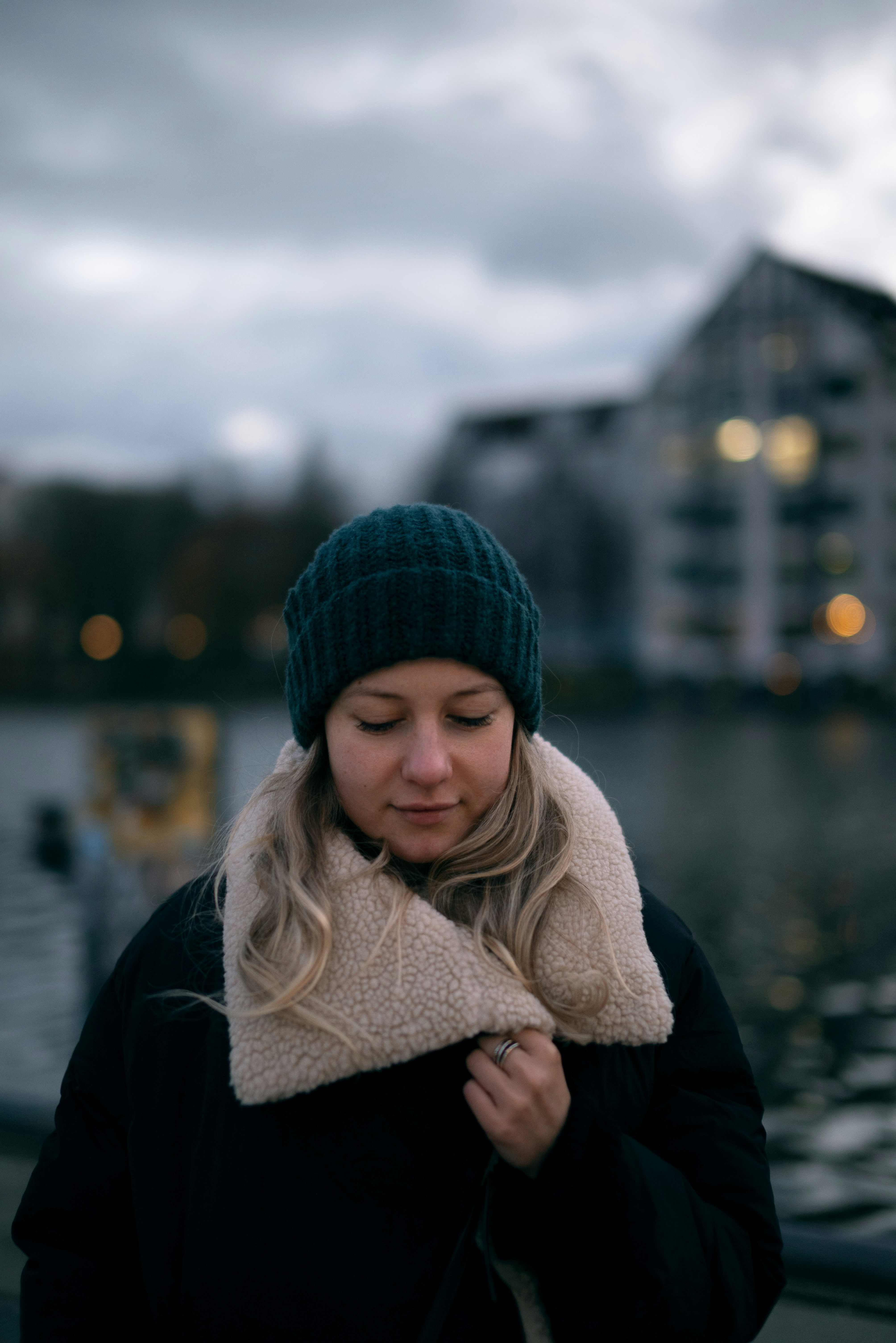 Woman in a cozy winter outfit gazes downward, with a blurred waterfront backdrop and soft bokeh lights illuminating the scene.