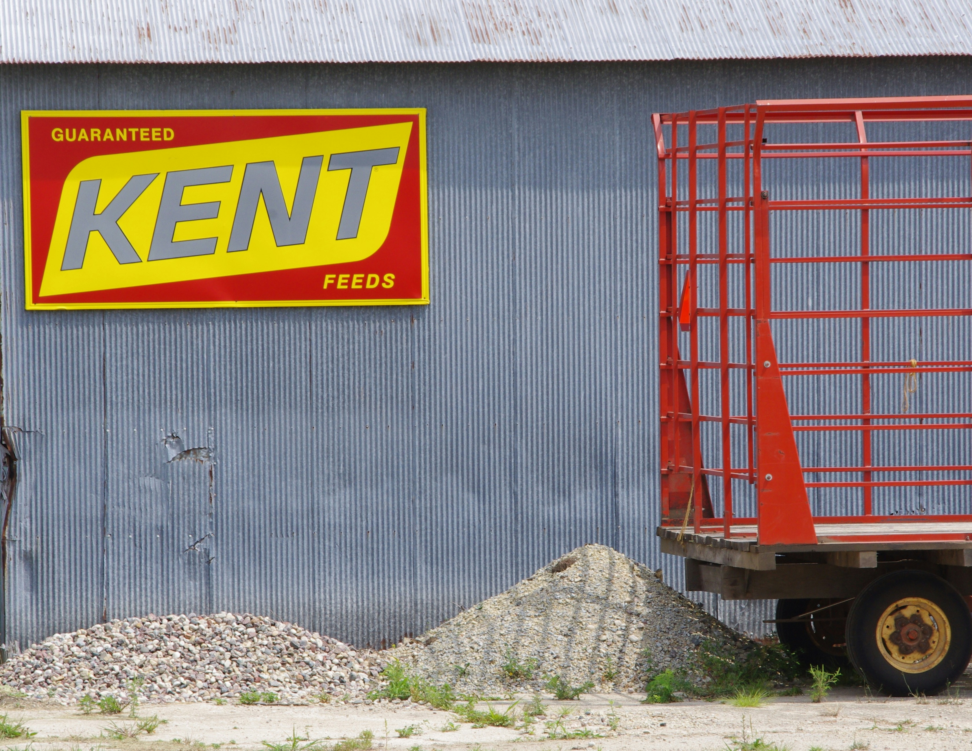 Vibrant Kent Feeds sign against a weathered blue wall, complemented by a red cart and gravel pile, showcasing a blend of industry and rural charm.