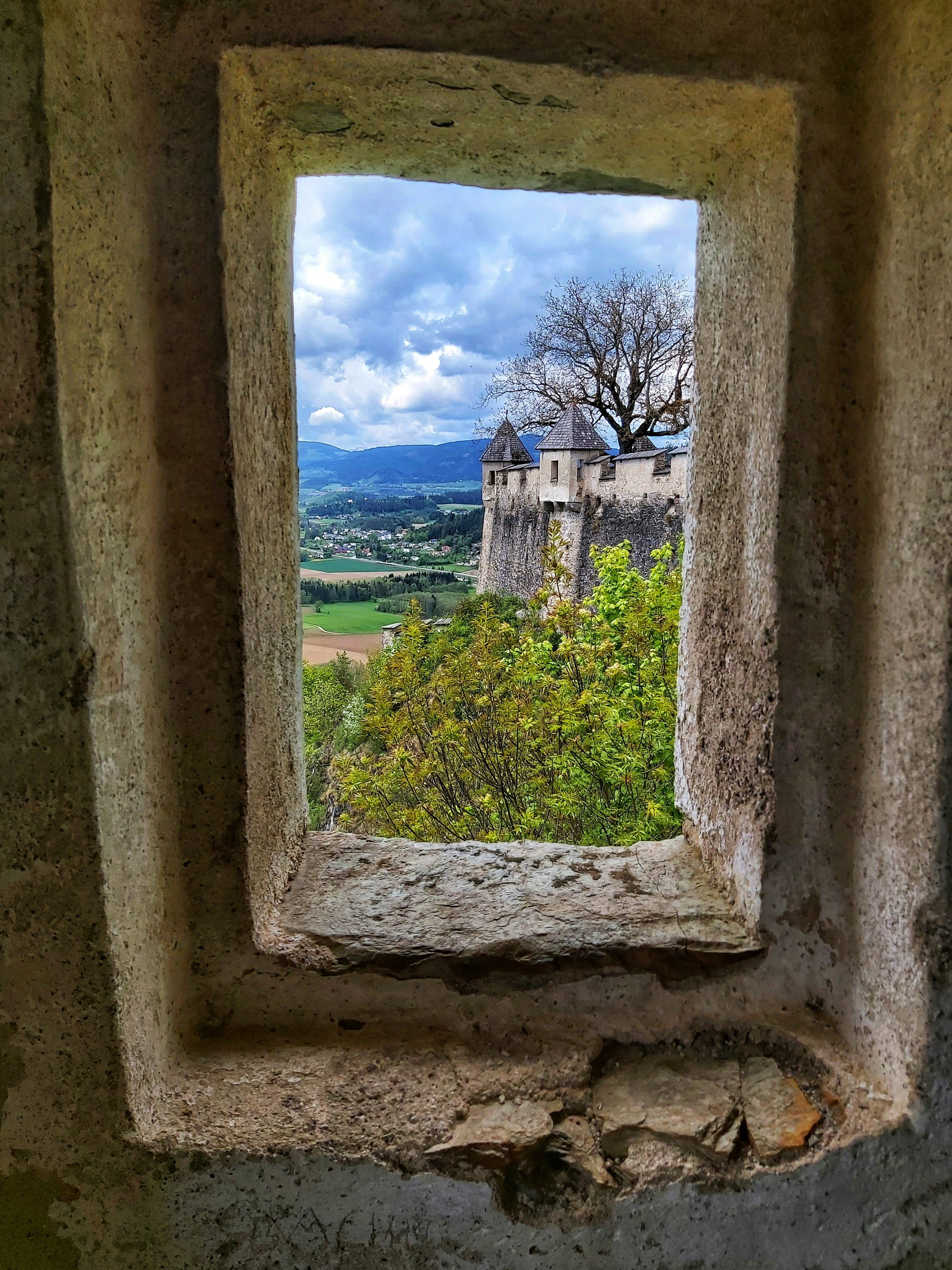 View of a historic castle framed by a stone window, showcasing lush greenery and rolling hills under a cloudy sky.
