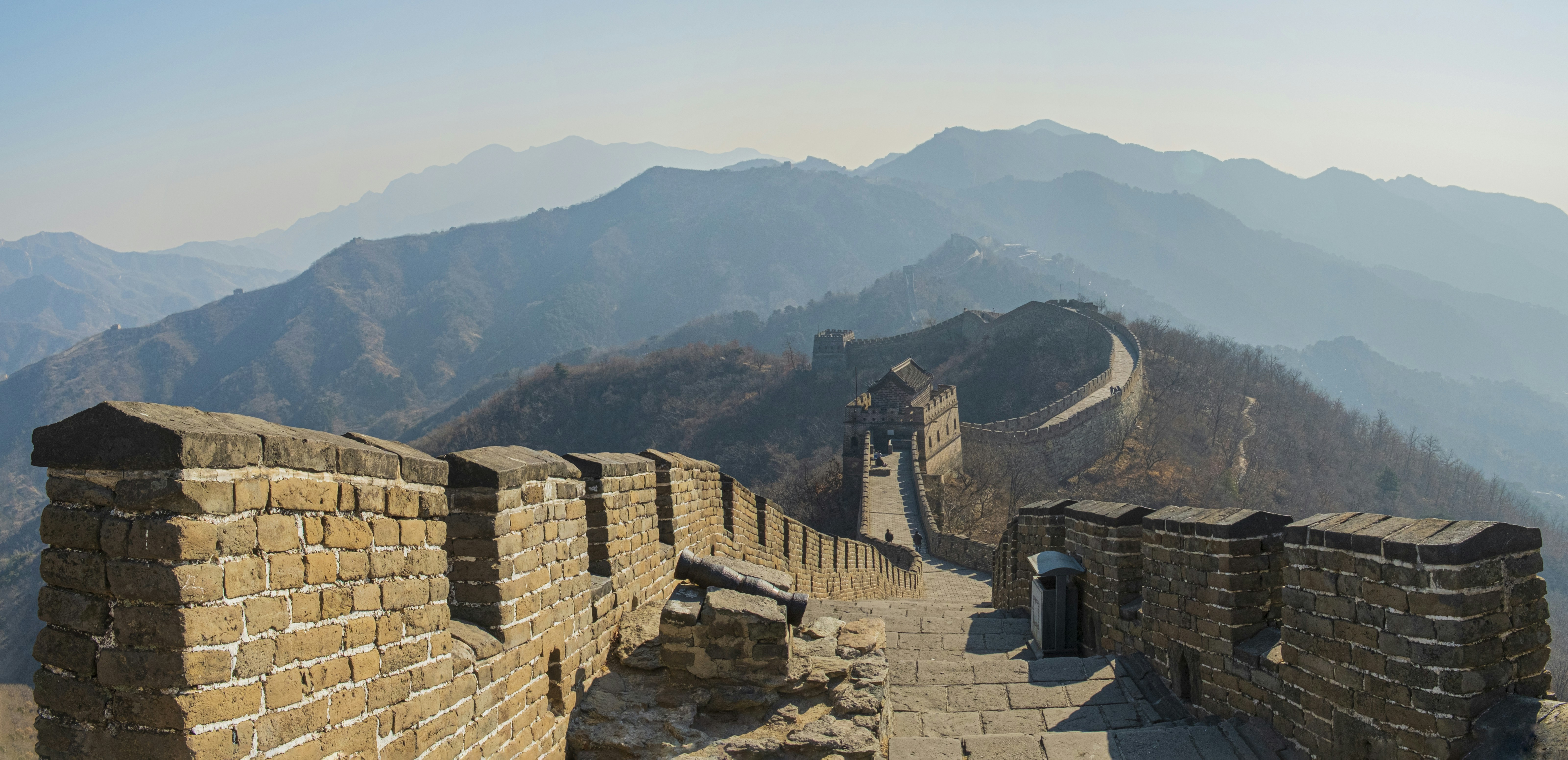 Ancient stone pathway of the Great Wall winding through mountainous terrain, showcasing historical architecture against a hazy backdrop.