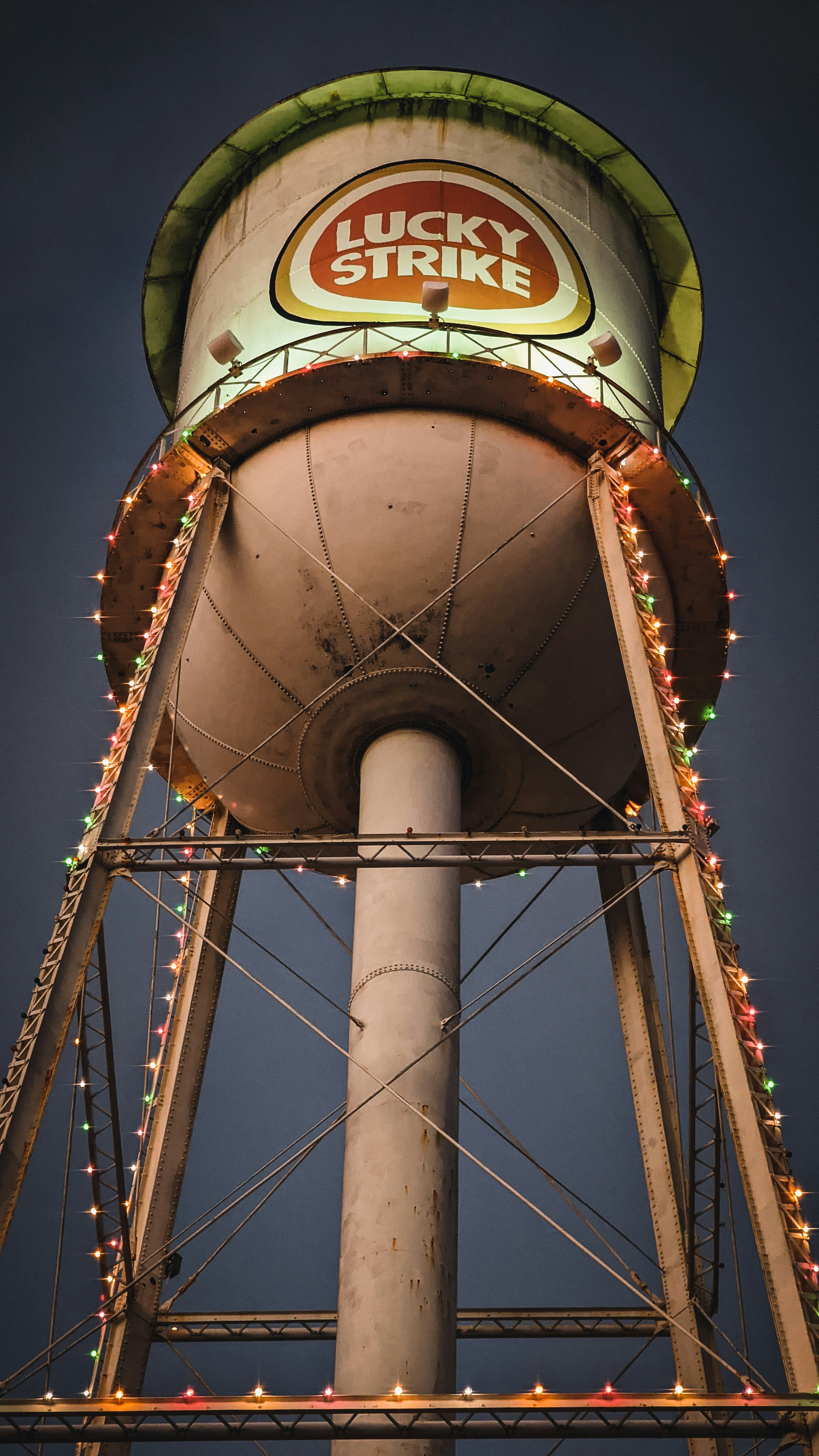 Night shot of a tall water tower wrapped in colorful string lights, crowned by a circular Lucky Strike logo.