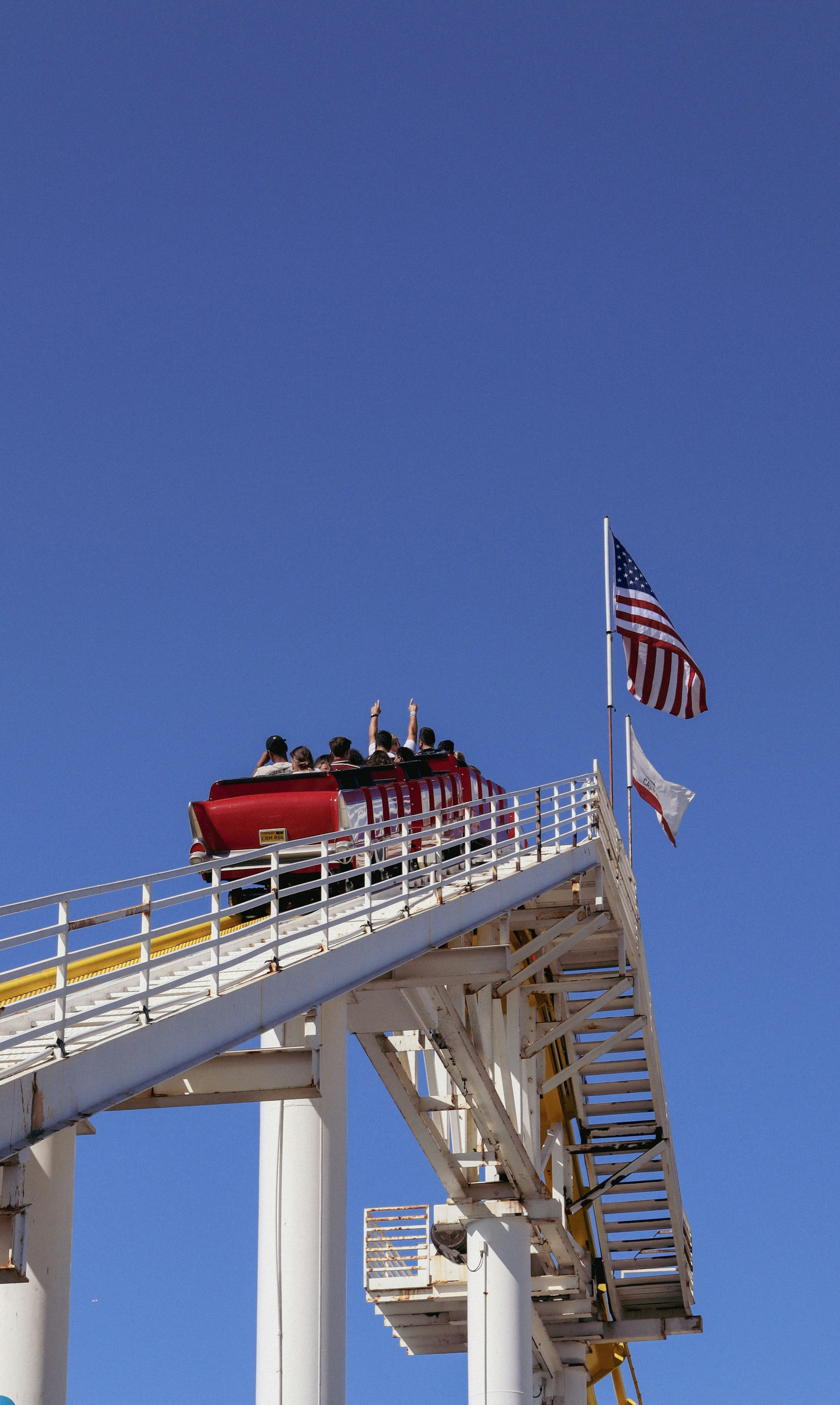 Roller coaster cart nearing the peak of a steep ascent, with riders raising their hands in excitement against a clear blue sky.