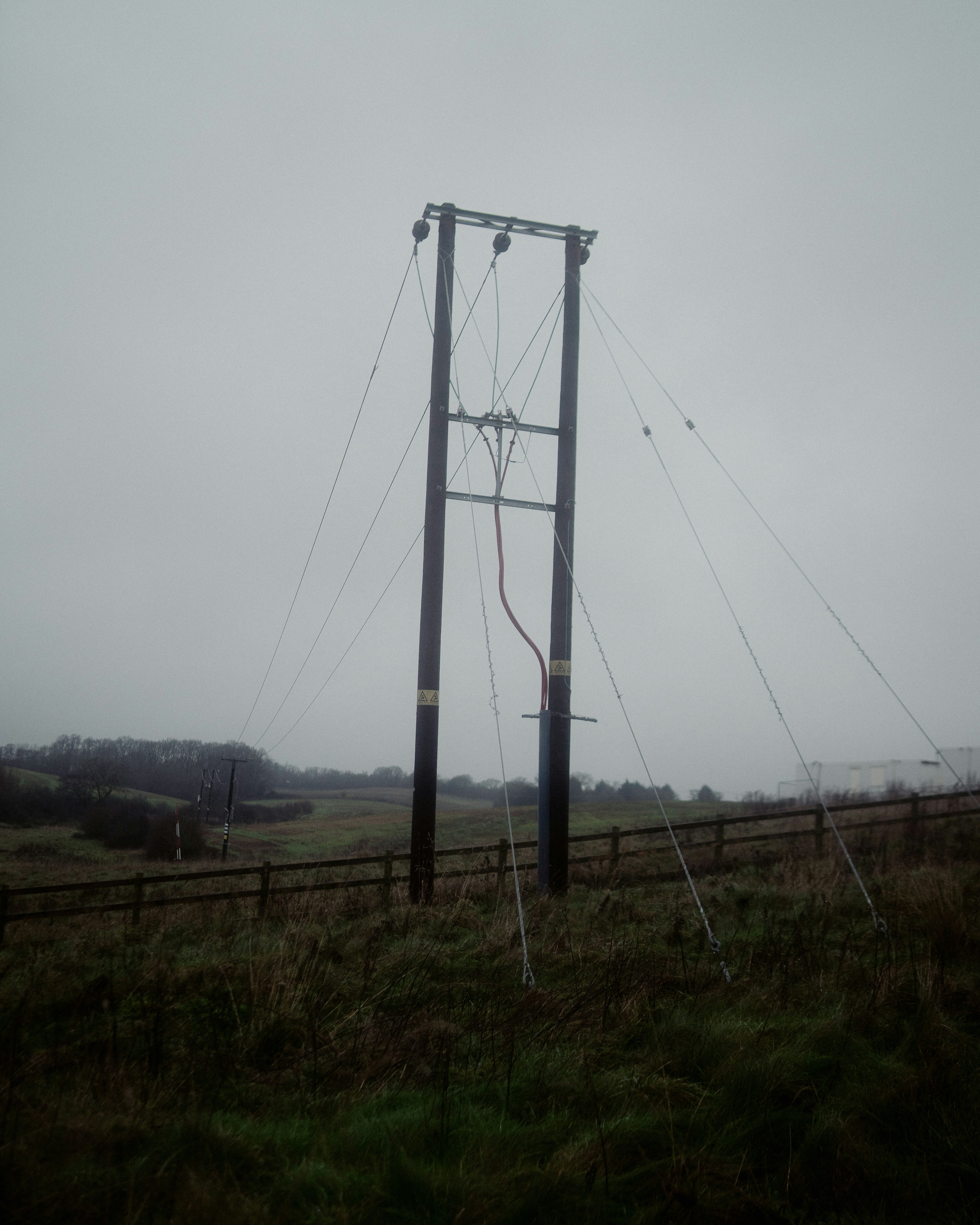 A tall power pole stands against a gray, overcast sky, surrounded by rolling hills and sparse vegetation. The scene evokes a sense of isolation and industrial presence in nature.