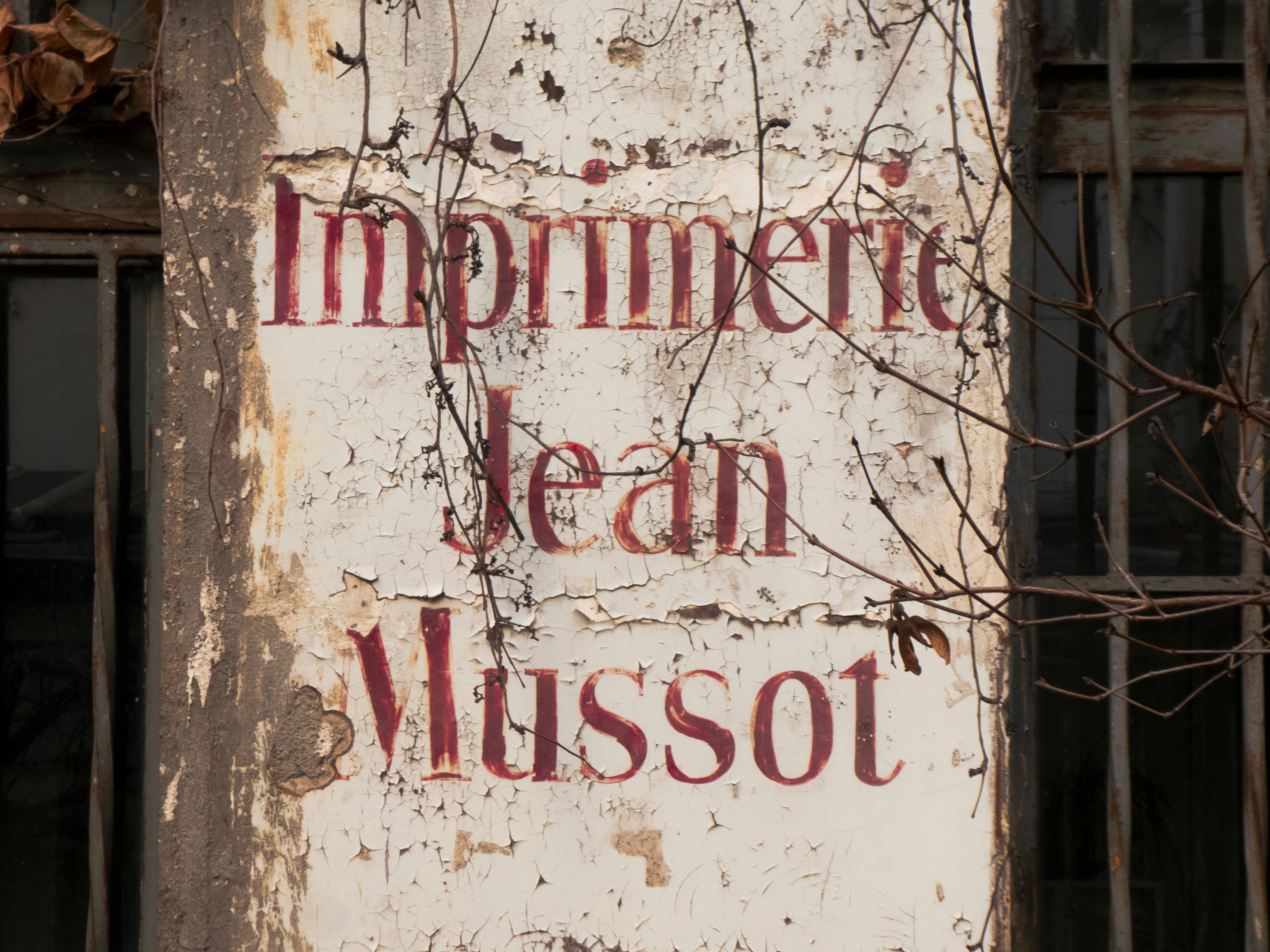 Faded signage of 'Imprimerie Jean Vlussot' on a weathered wall, surrounded by creeping vines. The peeling paint adds a nostalgic touch.