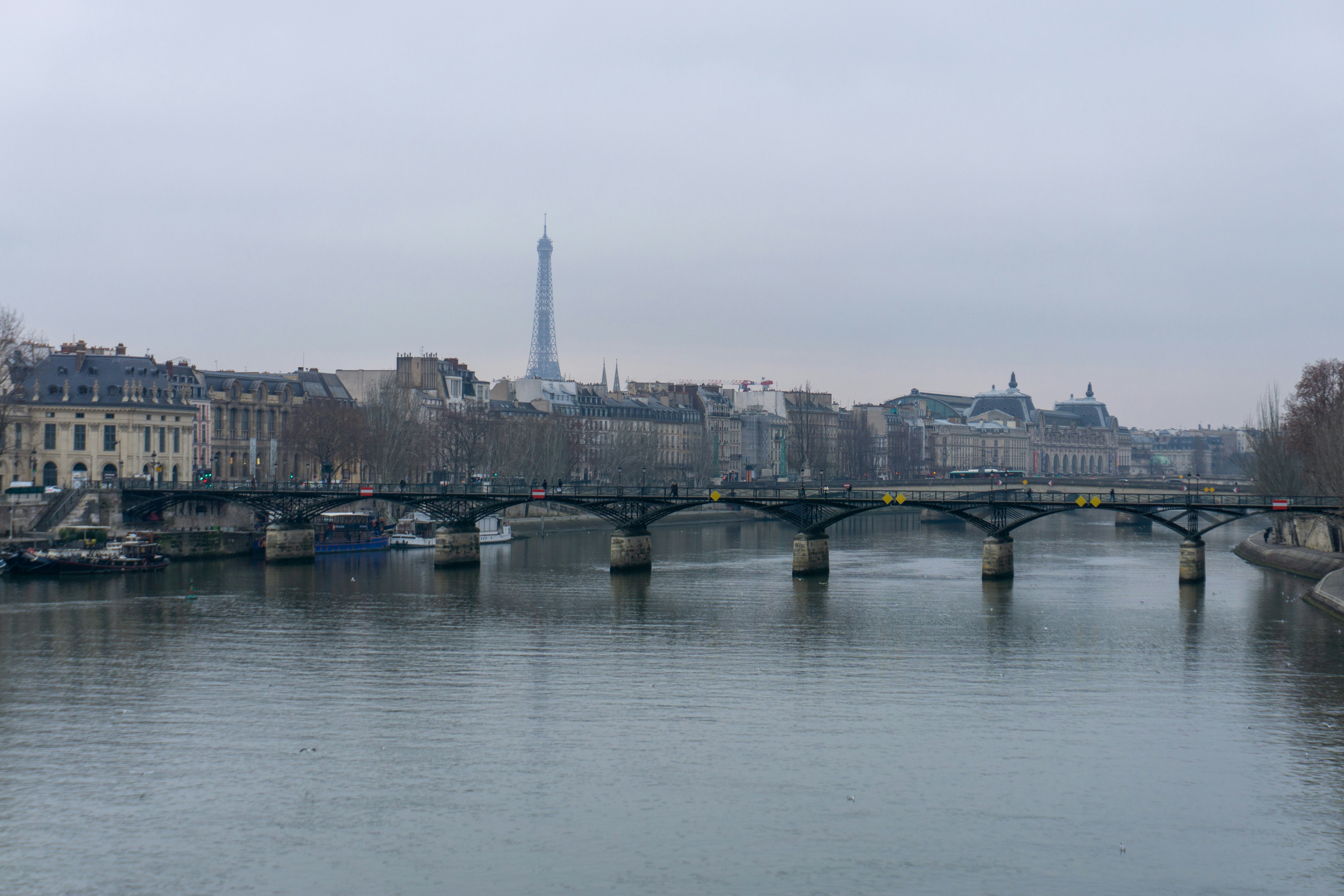 A tranquil view of the Seine River with a bridge in the foreground and the Eiffel Tower rising in the background, enveloped in a soft, overcast sky.