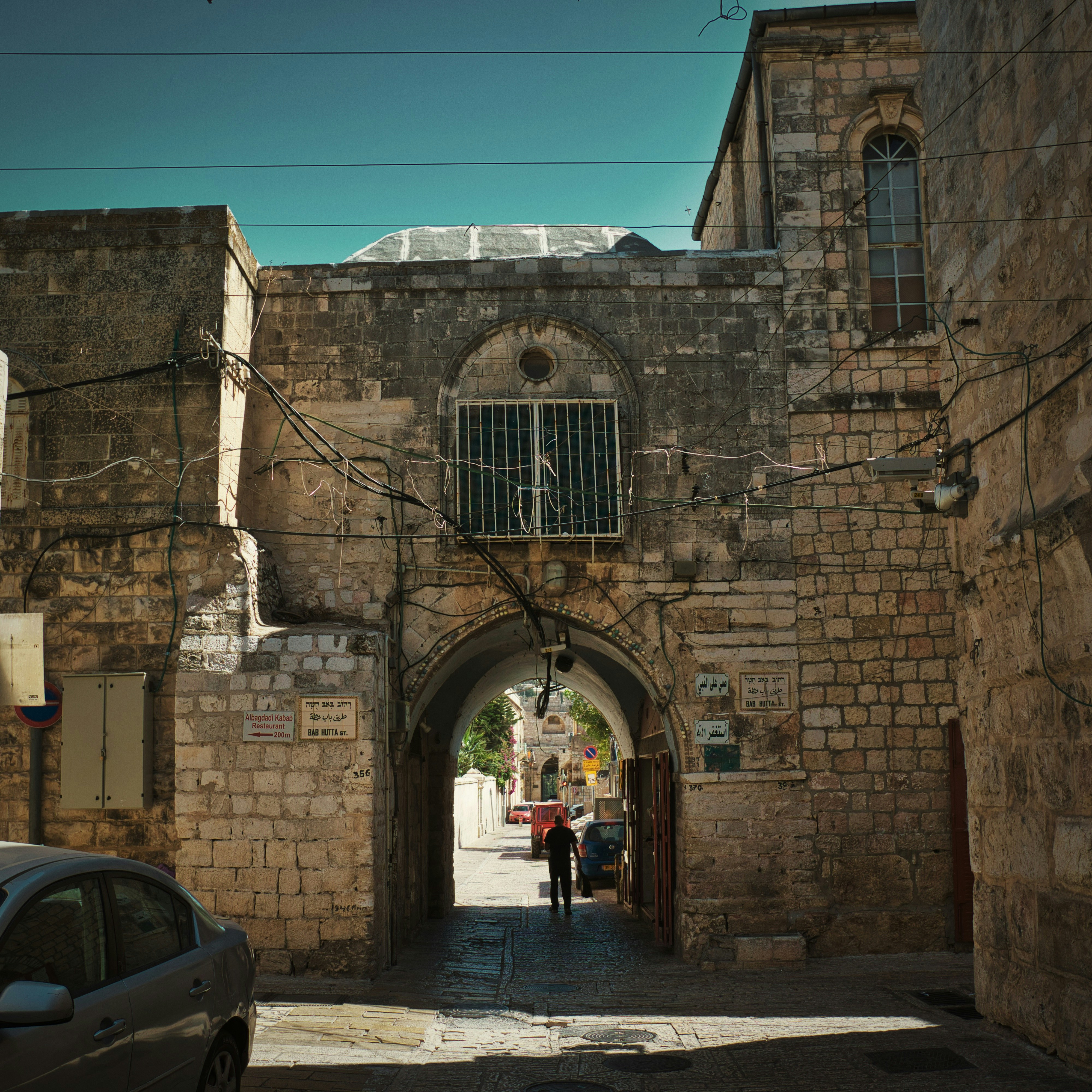 Historic stone archway leading to a vibrant street, framed by textured walls and overhead wires. A figure walks towards the entrance.