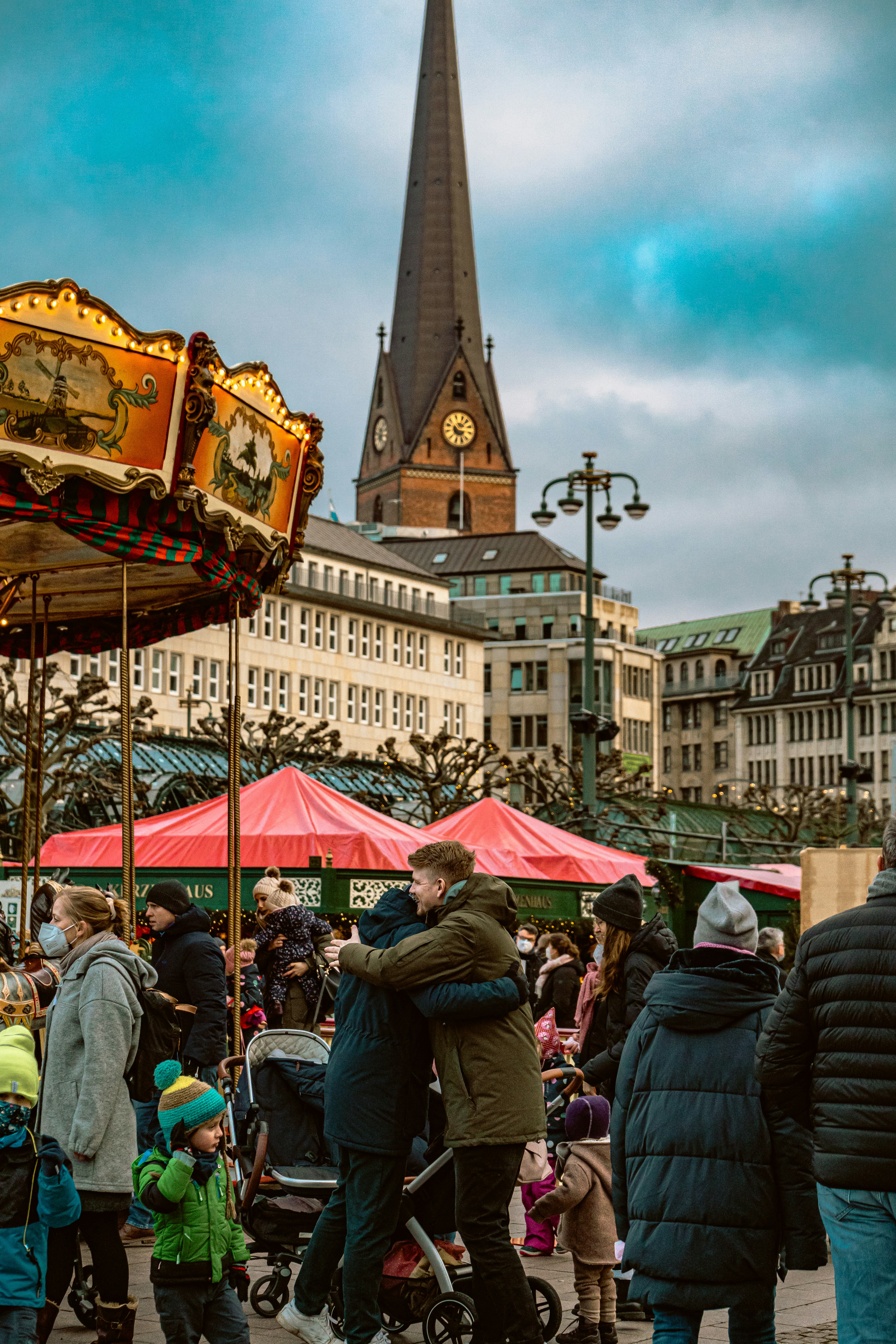 A festive scene captures families enjoying a carousel in a bustling square, with a prominent clock tower in the background. The atmosphere is lively and filled with warmth.