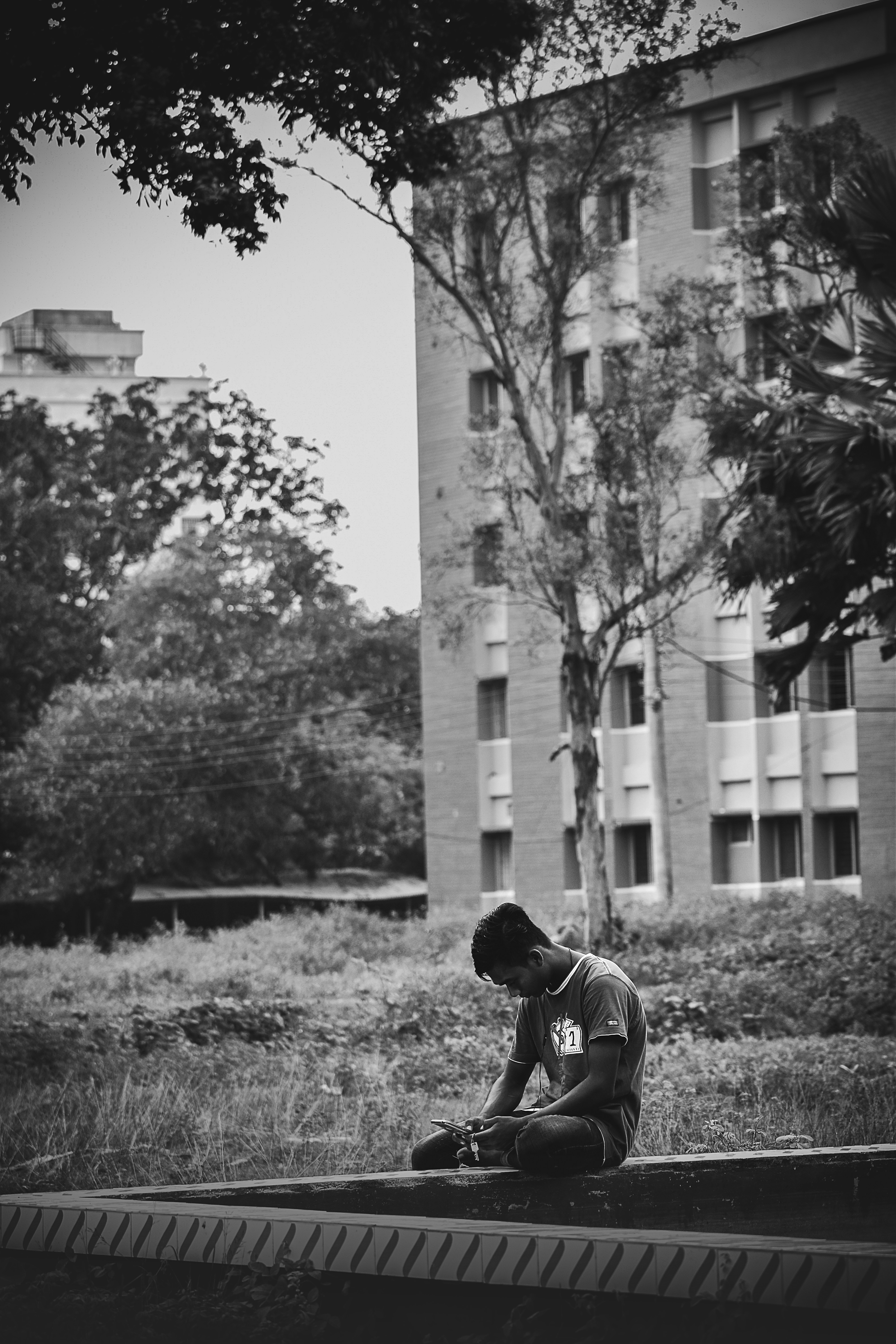 A young man sits pensively on a ledge, surrounded by lush greenery and urban architecture, embodying a moment of introspection. The scene is rendered in black and white, enhancing the mood of reflection.