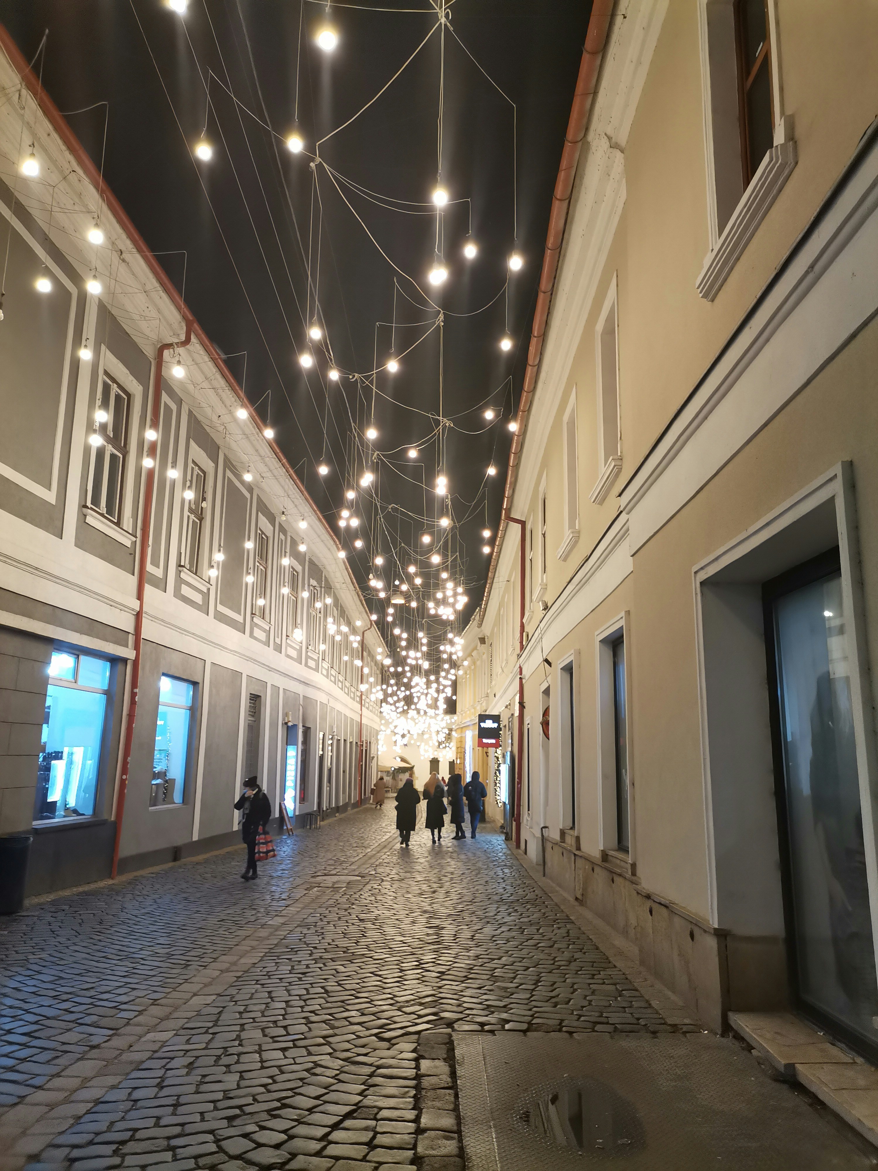 Charming cobblestone street illuminated by hanging lights, with people walking in the distance. The warm glow creates a cozy atmosphere.