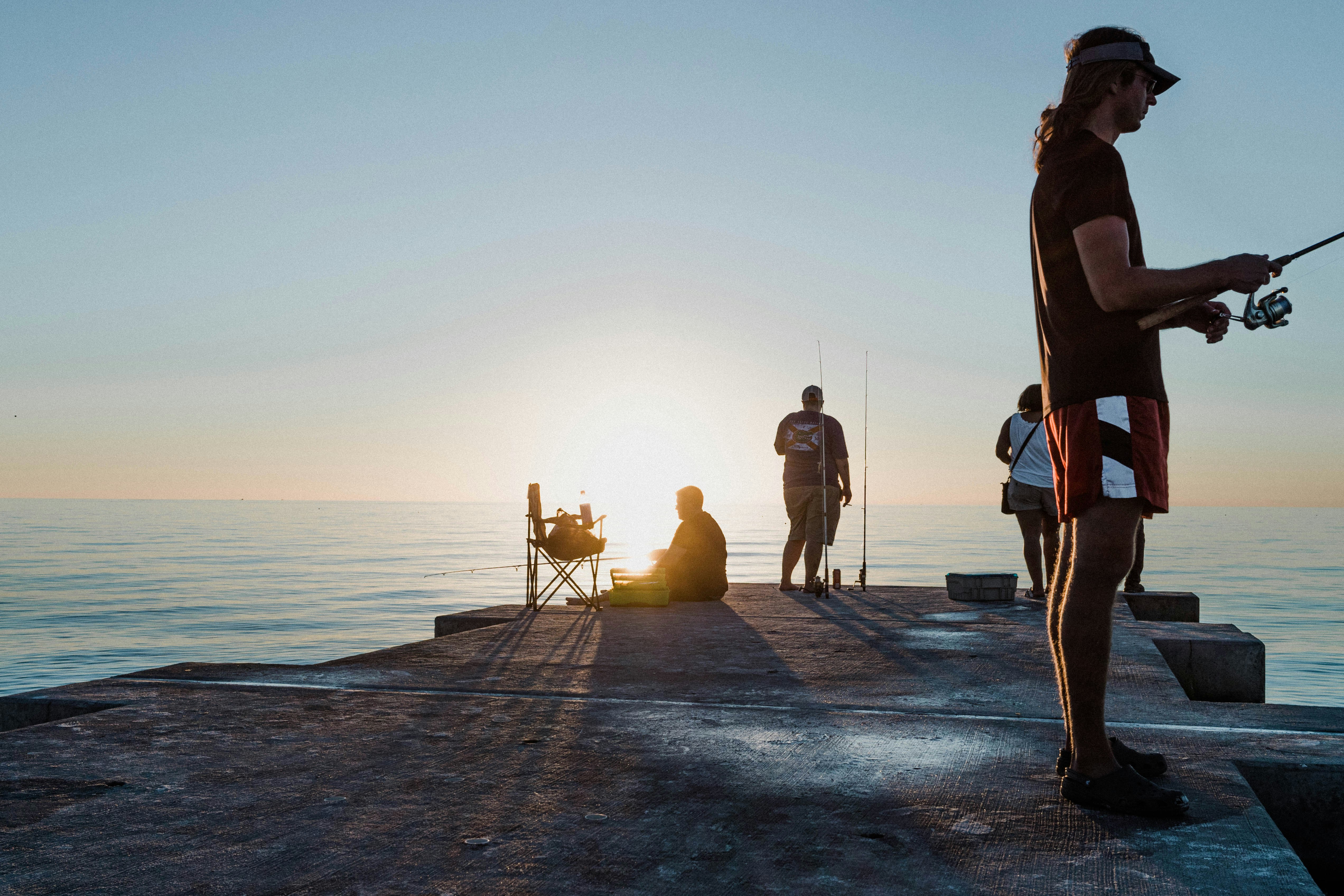 Silhouetted figures fishing on a pier at sunset with calm ocean waters and a glowing horizon.