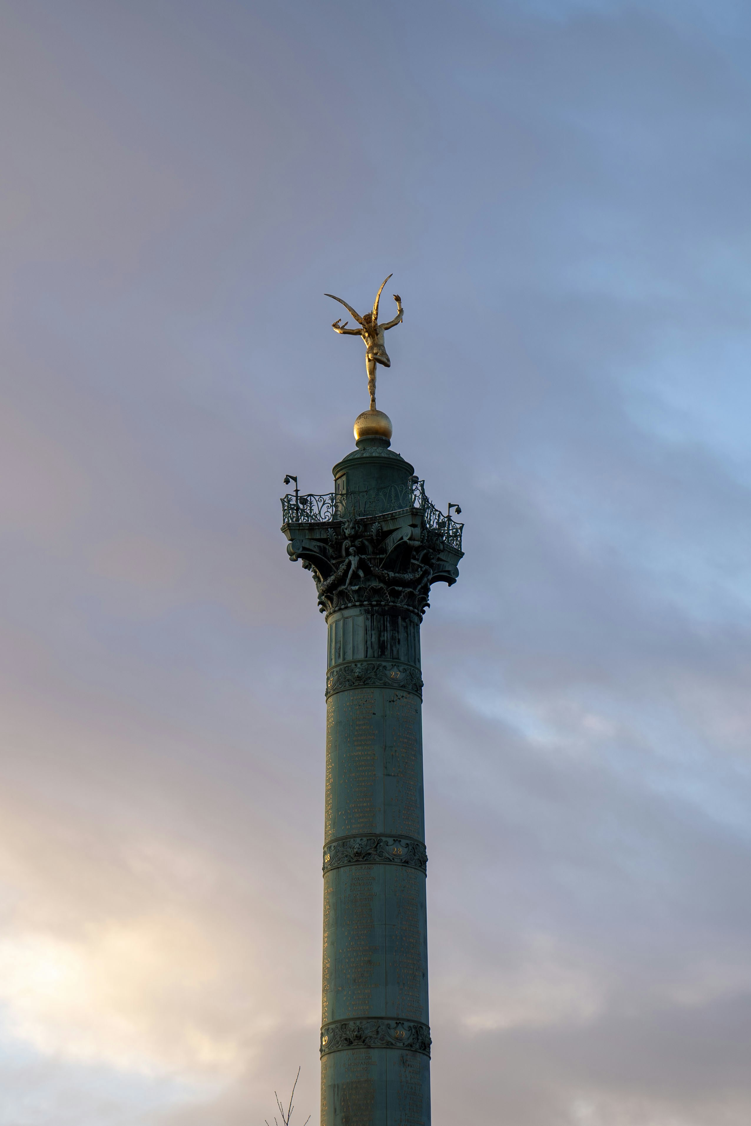 A majestic column topped with a golden figure, representing victory, stands against a backdrop of soft pastel clouds at dusk.