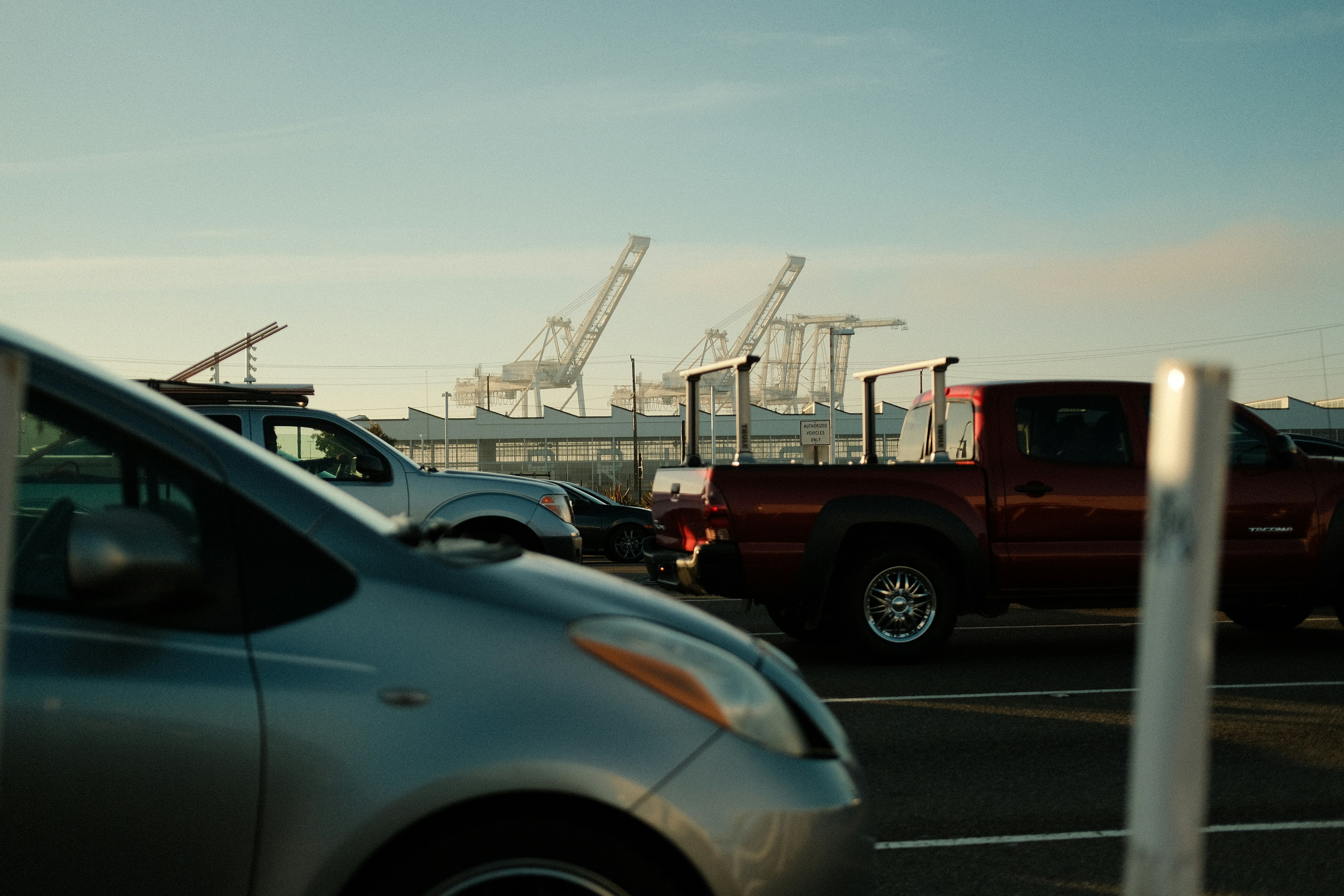 Red pickup truck and silver car parked with cranes visible in the background under a clear sky.